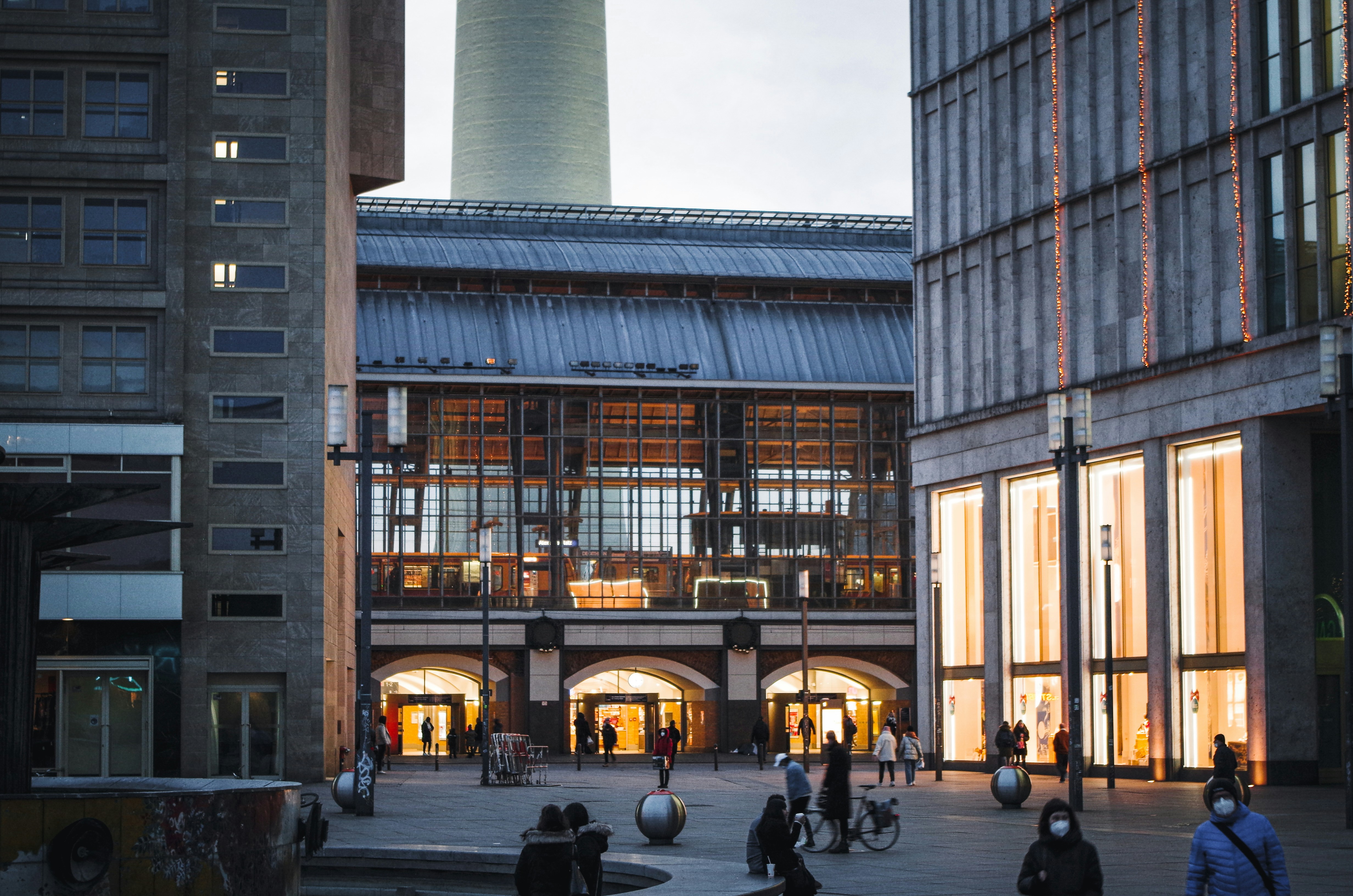 Modern architectural elements frame a bustling plaza, illuminated storefronts contrasting with the twilight sky. A prominent chimney rises in the background.