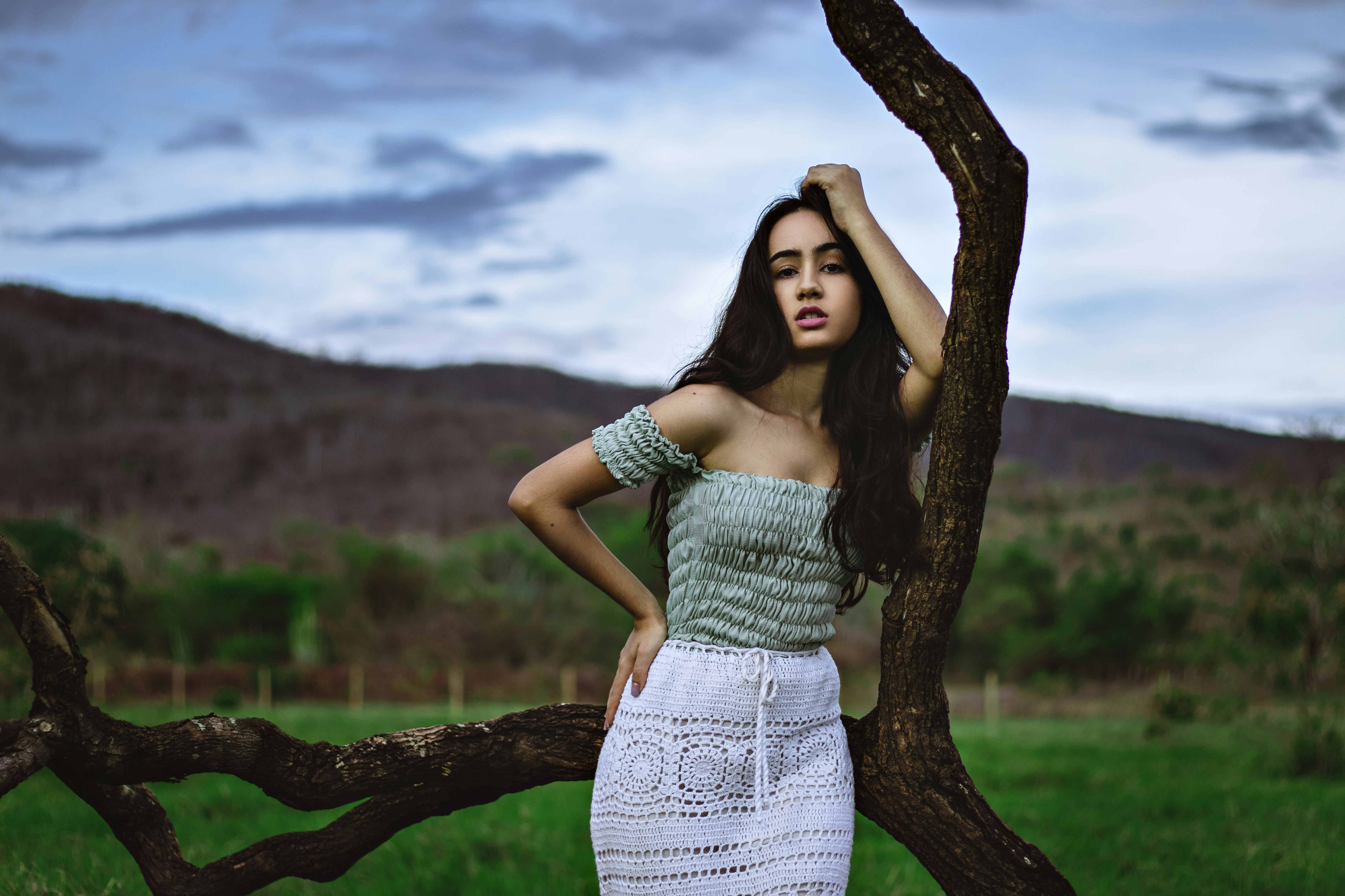A young woman poses gracefully against a twisted tree branch, showcasing a blend of natural beauty and elegance in a lush green landscape.