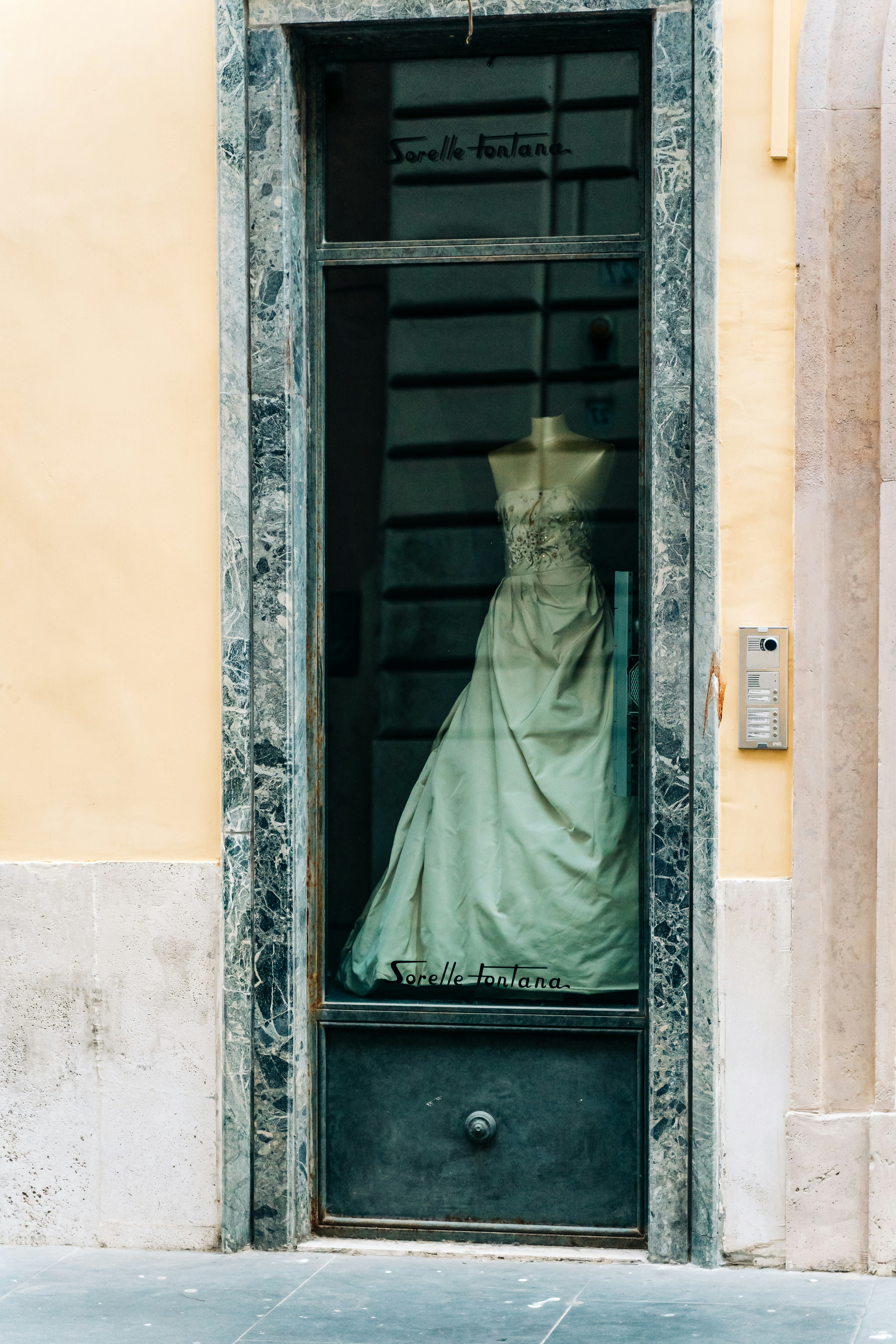 Elegant white wedding dress displayed in a tall, narrow window with ornate stone framing.