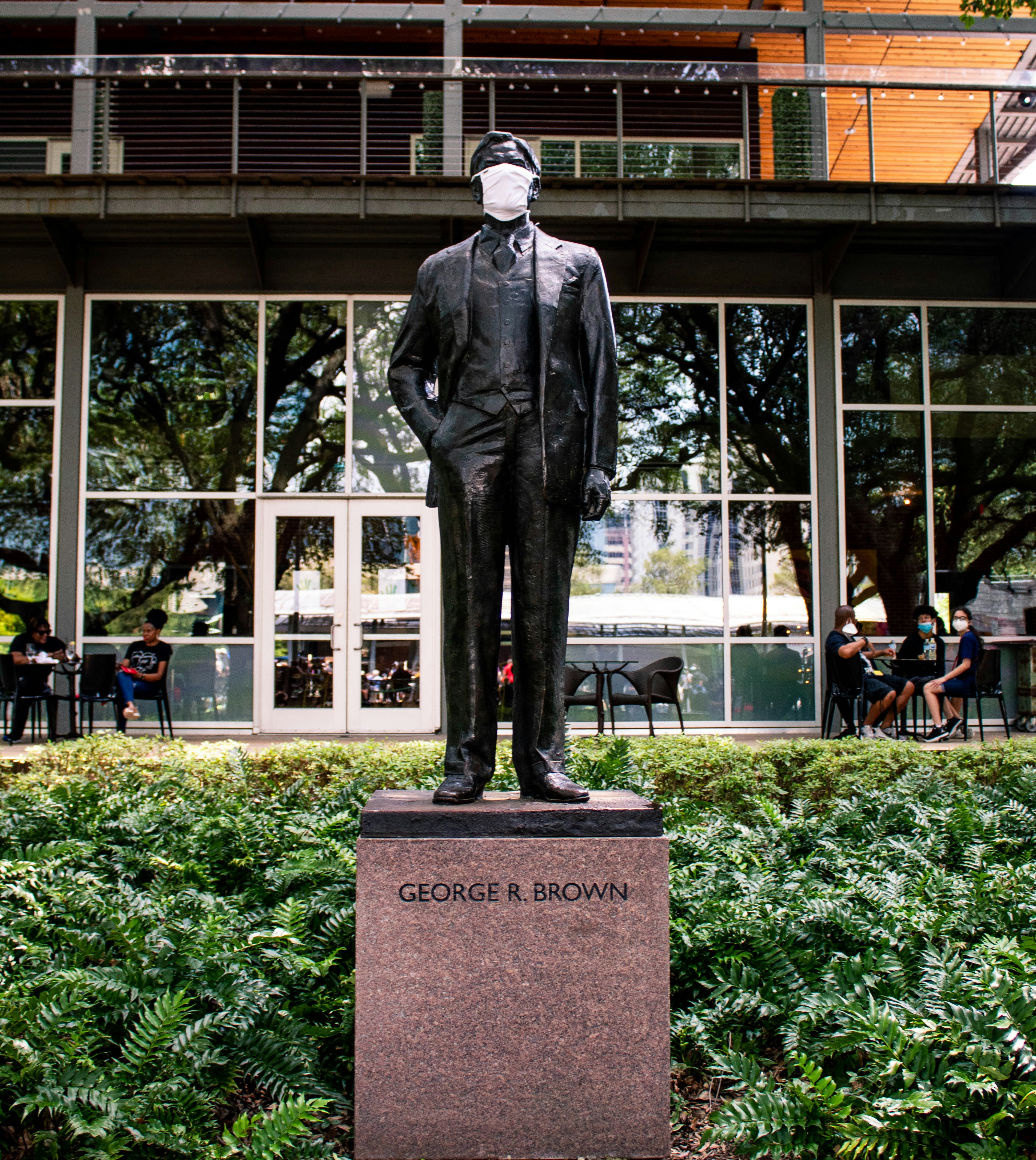 Bronze statue of George R. Brown, adorned with a mask, stands amidst lush greenery outside a contemporary building.