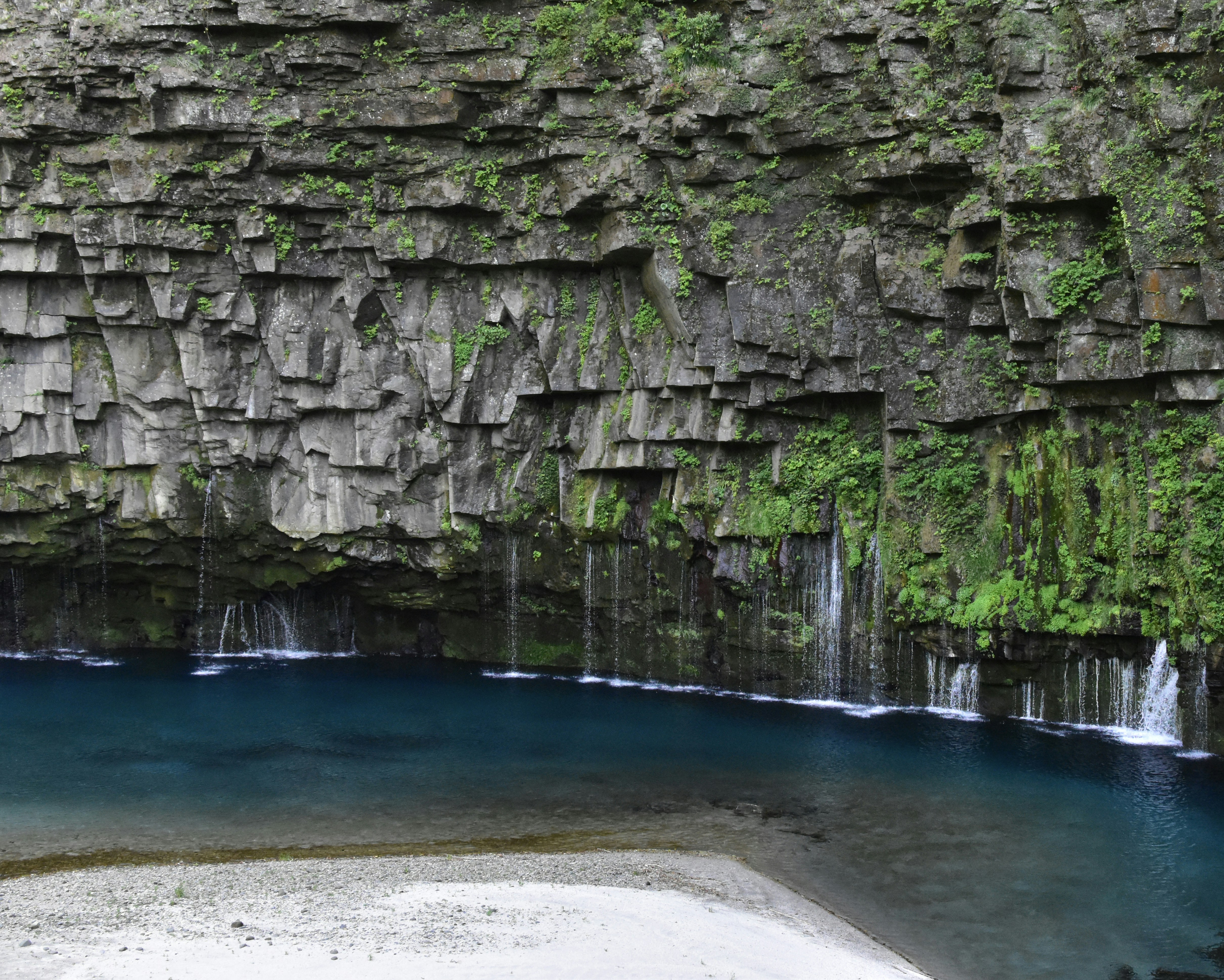 Small waterfalls trickle down a lush, green-clad basalt cliff into a serene blue pool.