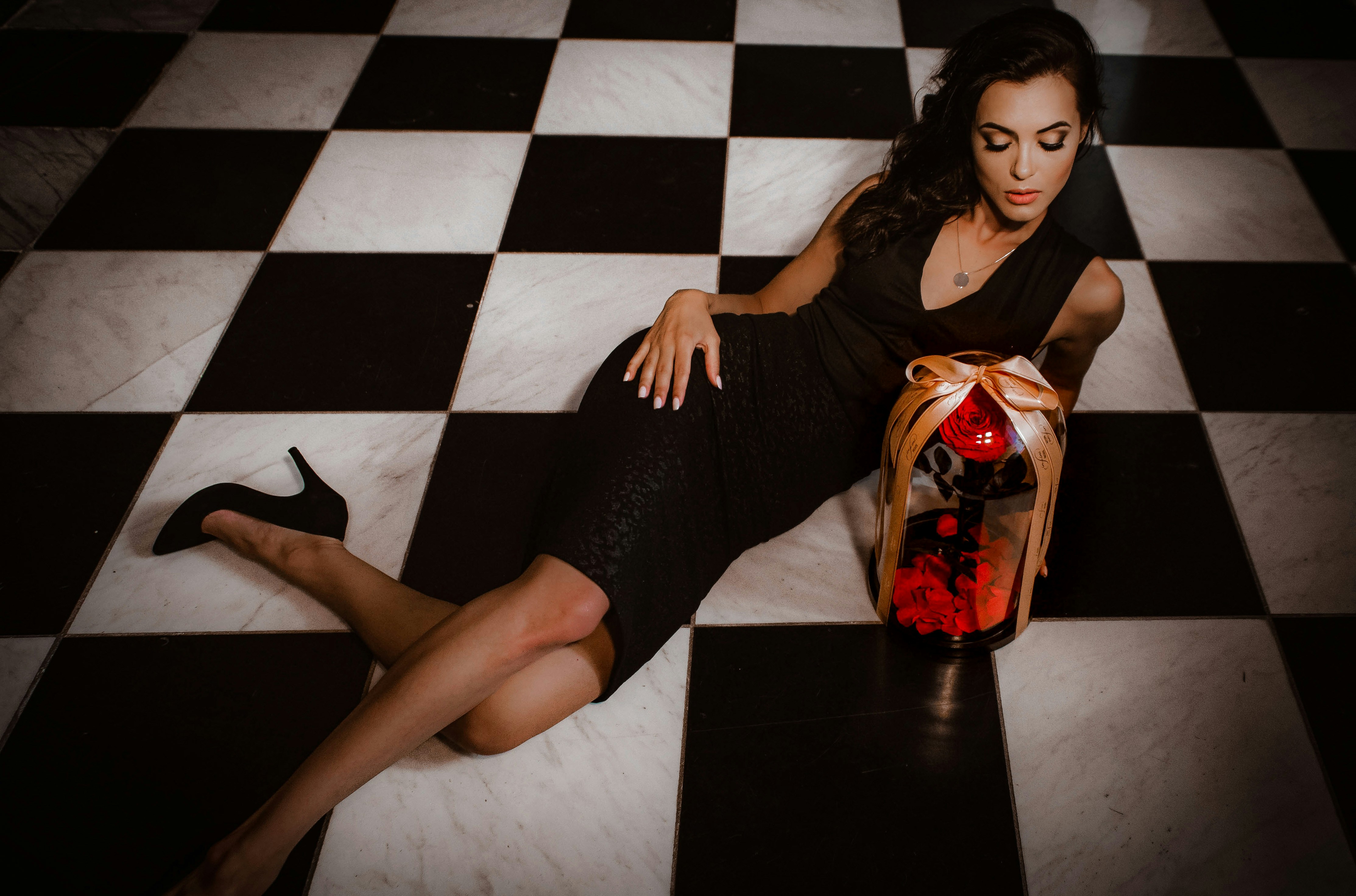 Woman in a black dress reclines on a black-and-white checkered floor with a red rose encased in glass.