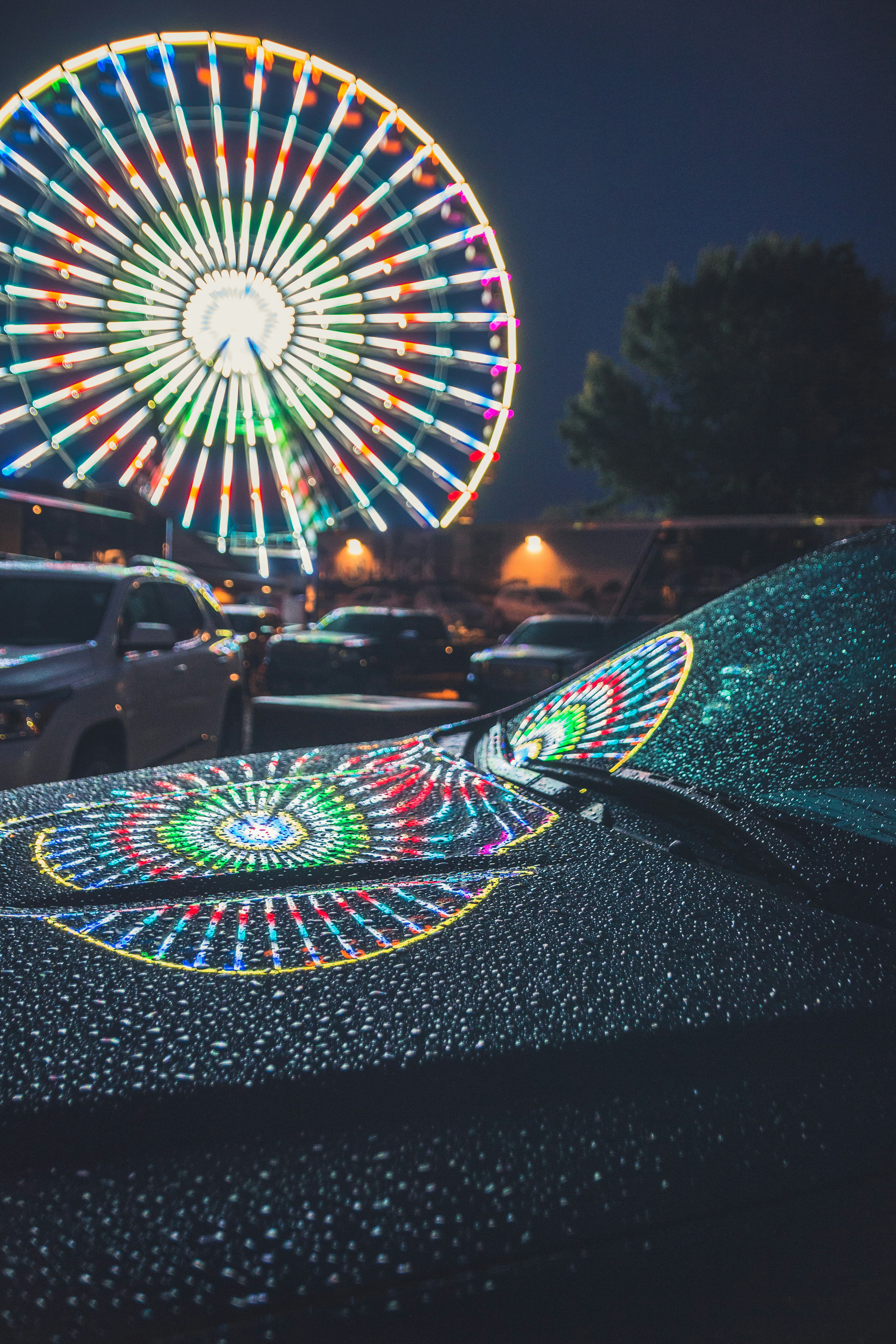 Vibrant reflections of a Ferris wheel's colorful lights dance on a rain-soaked car hood, creating a mesmerizing interplay of color and texture.