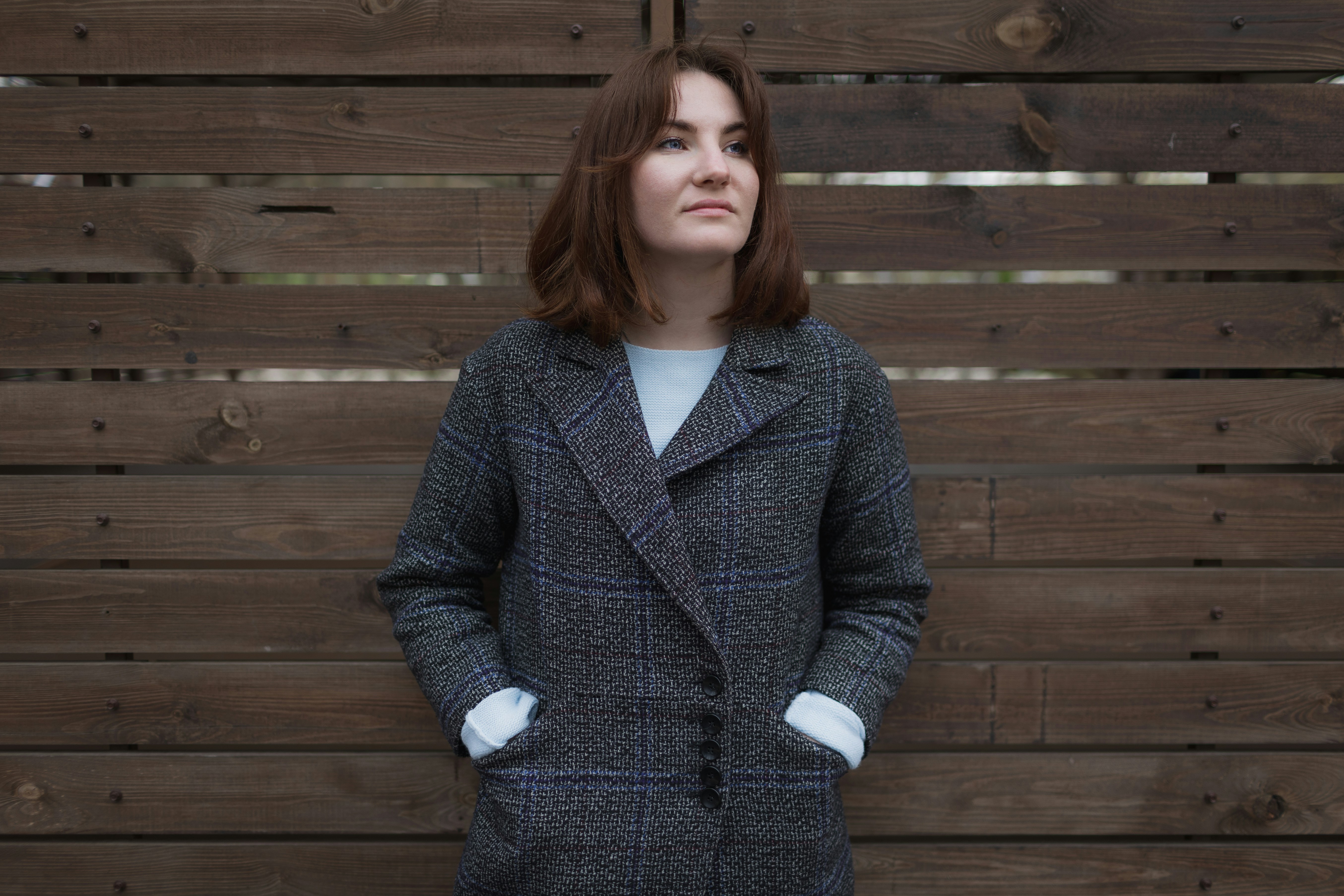 Woman in a plaid coat stands against a wooden fence, hands in pockets.