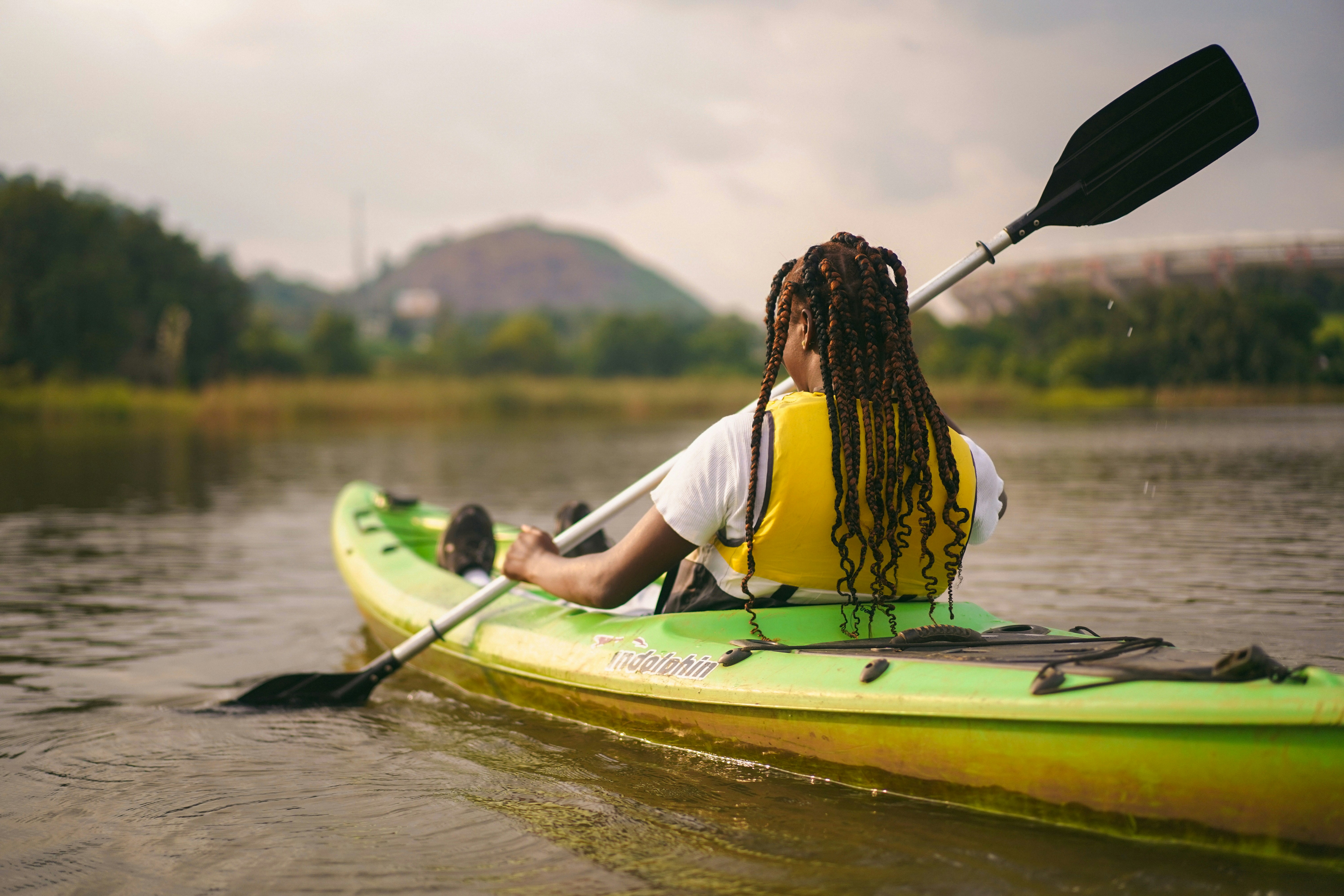 Person in a yellow life jacket paddling a green kayak on a calm lake under a cloudy sky.