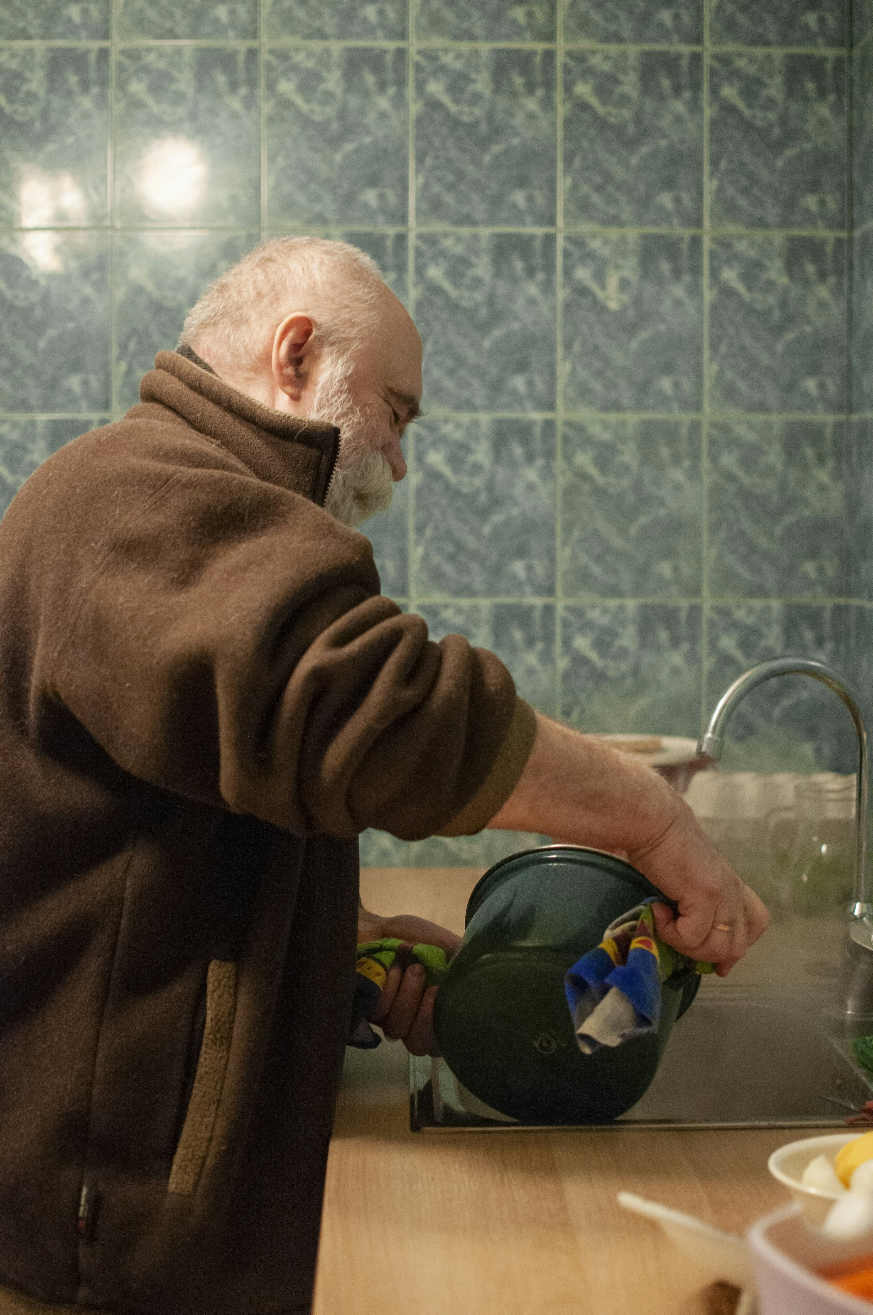 Elderly man pouring water from a green watering can into a sink, surrounded by a cozy kitchen atmosphere.