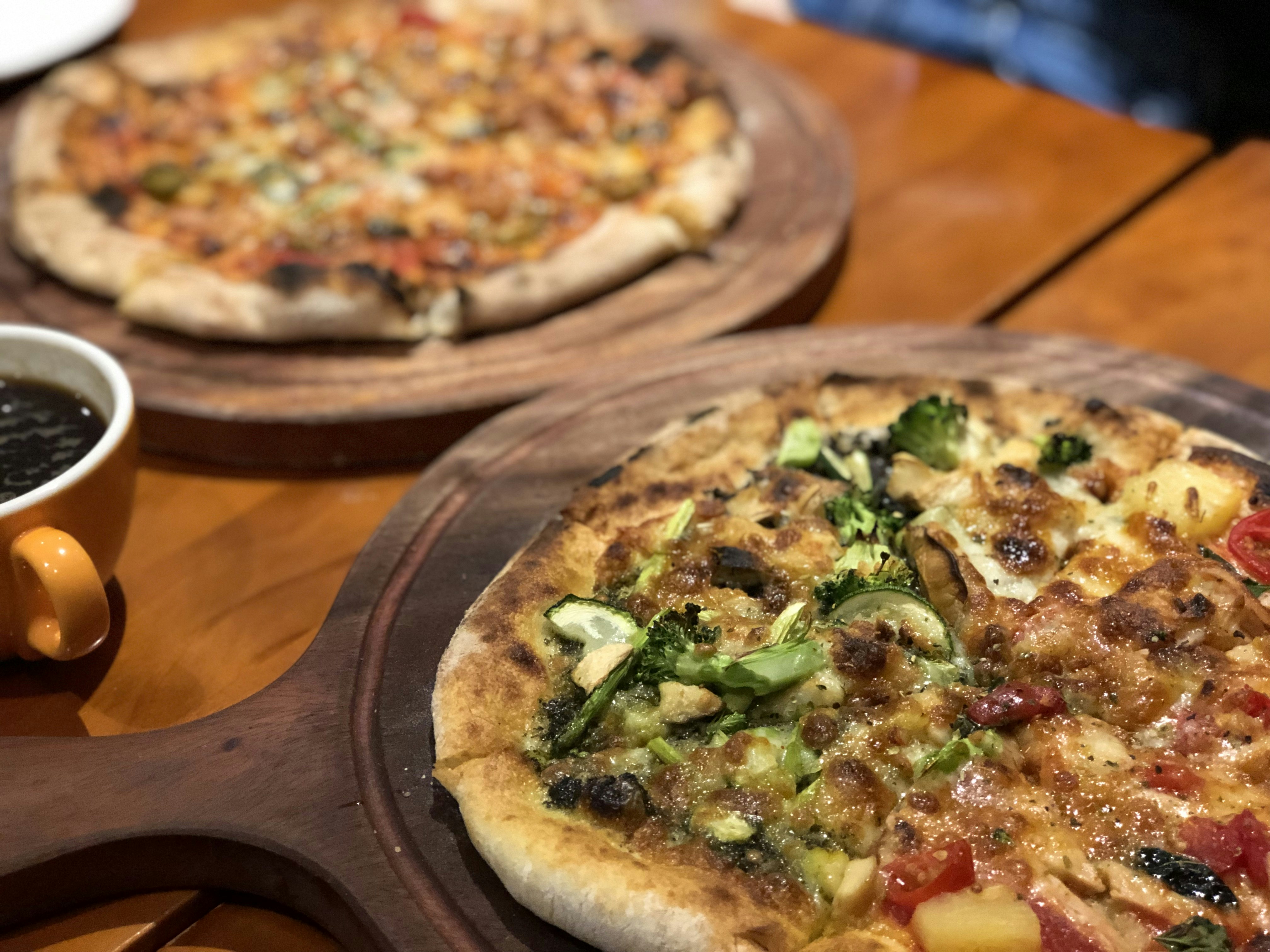 Two gourmet pizzas with varied toppings on wooden boards beside a cup of coffee.