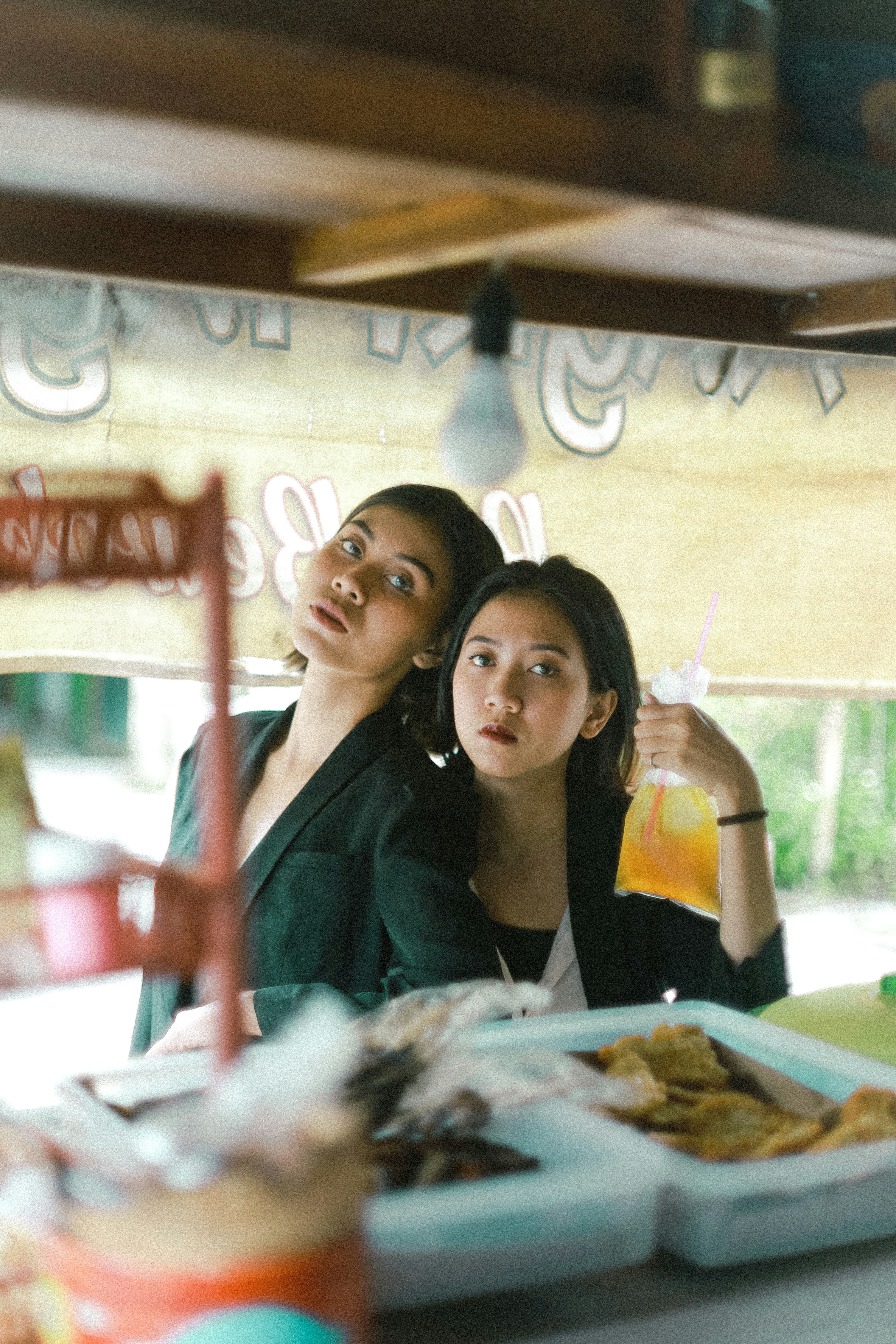 Two young women pose playfully at a vibrant food stall, one holding a drink while the other gazes thoughtfully. The scene captures a lively atmosphere filled with colors and textures.