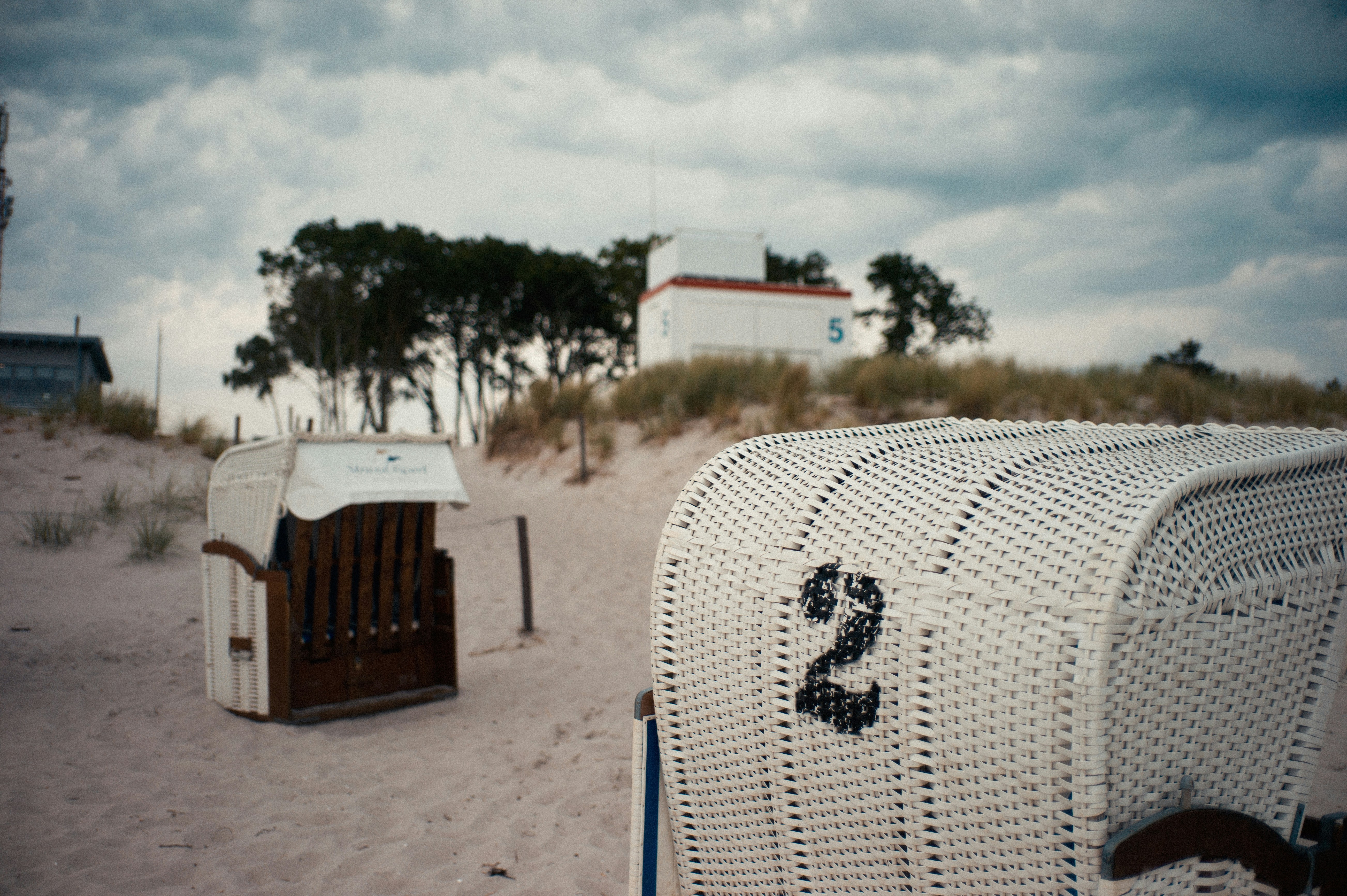 Woven beach chairs numbered 2 and 5 stand on sandy shore, framed by distant trees and a cloudy sky.