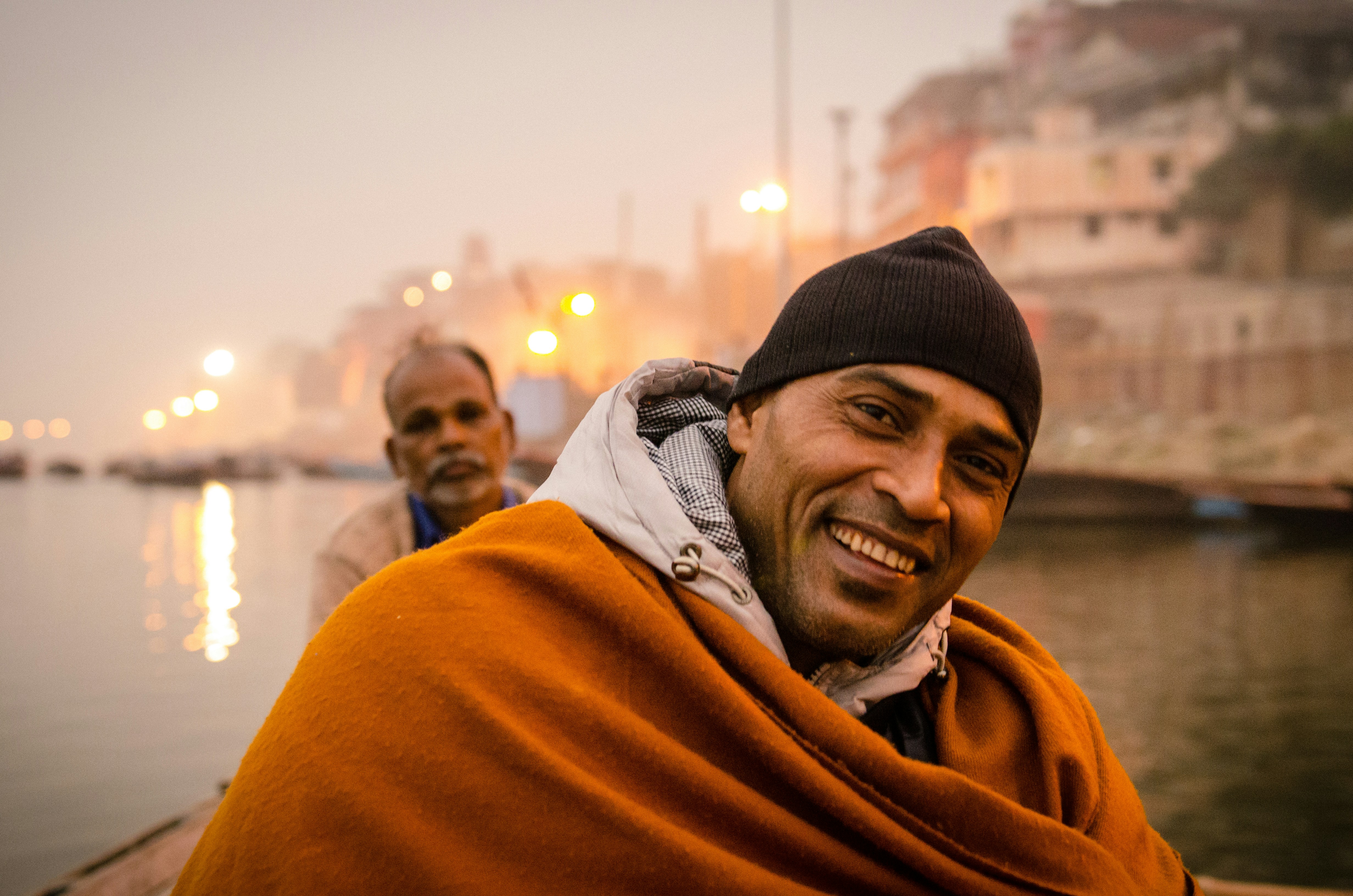 Two men in a boat on a tranquil river, one smiling warmly while wrapped in an orange blanket, with softly glowing lights in the background.
