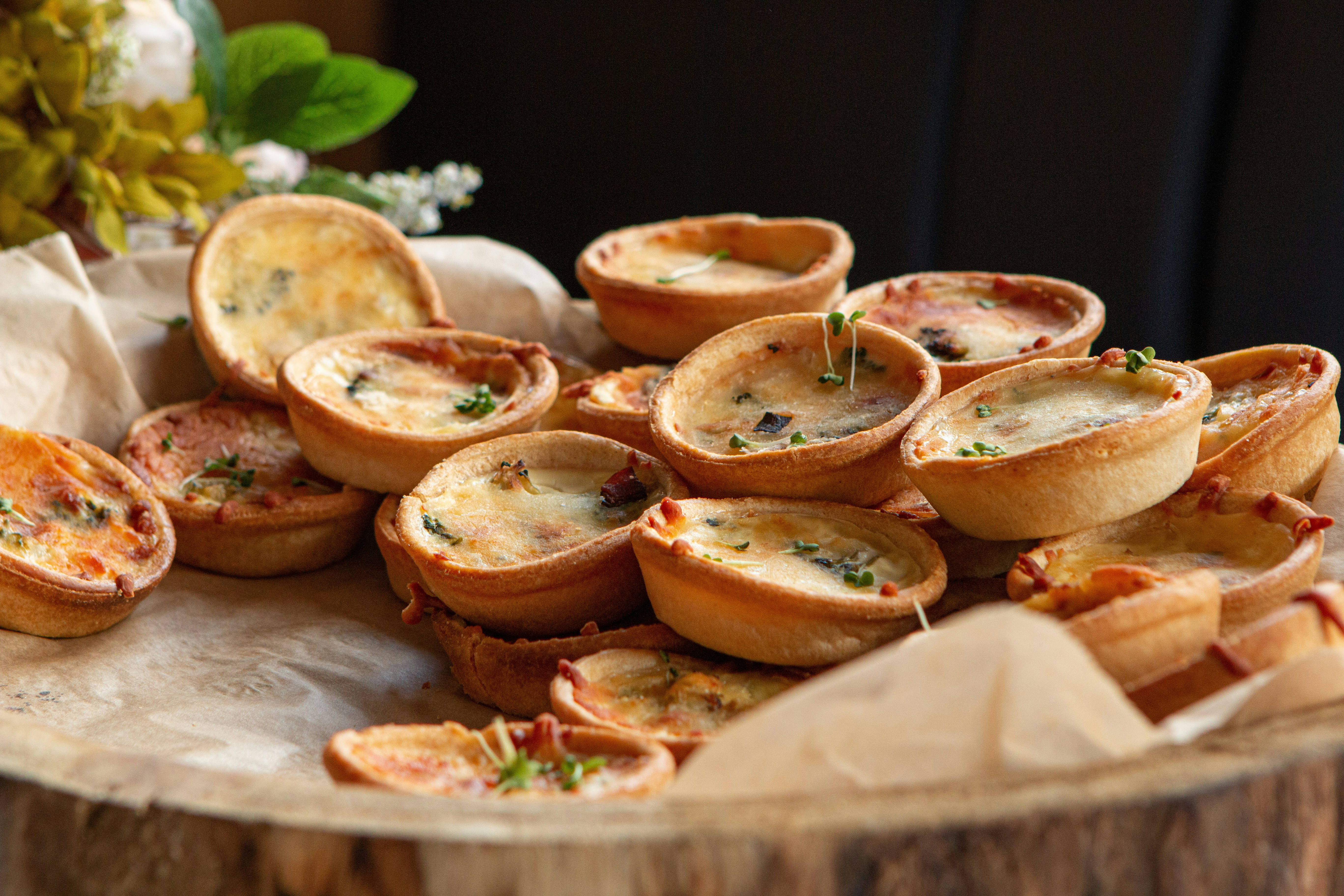 This image captures a selection of rustic mini quiches, artfully arranged on a wooden platter lined with parchment paper. The warm, golden hues of the pastry contrast beautifully with the vibrant greens of fresh herbs sprinkled on top. Soft, natural lighting enhances the inviting atmosphere, highlighting the textures and colors that make this culinary composition visually striking and appetizing.