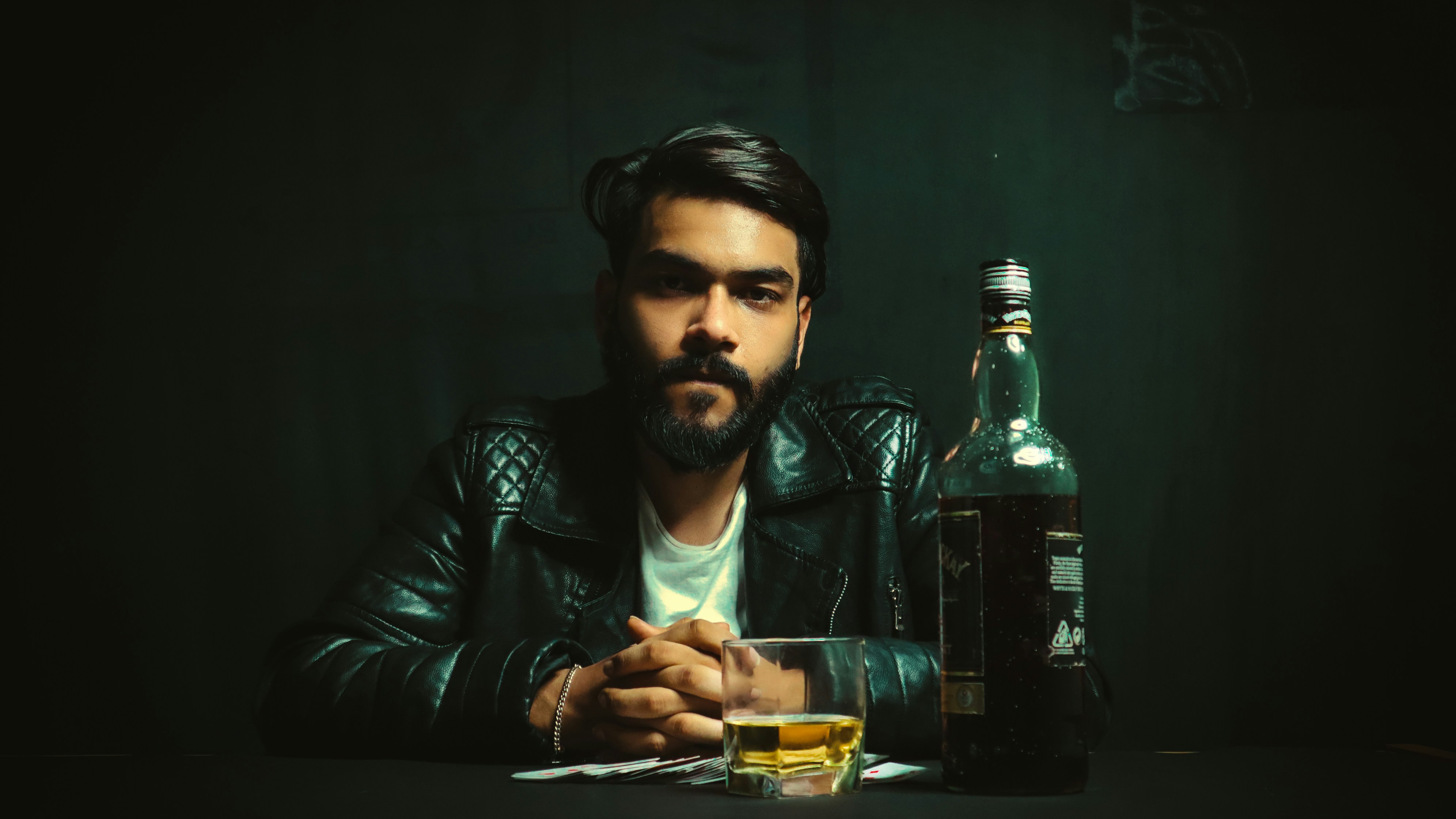 A brooding figure in a leather jacket sits at a table, flanked by a glass of whiskey and a bottle, enveloped in shadows and intrigue.