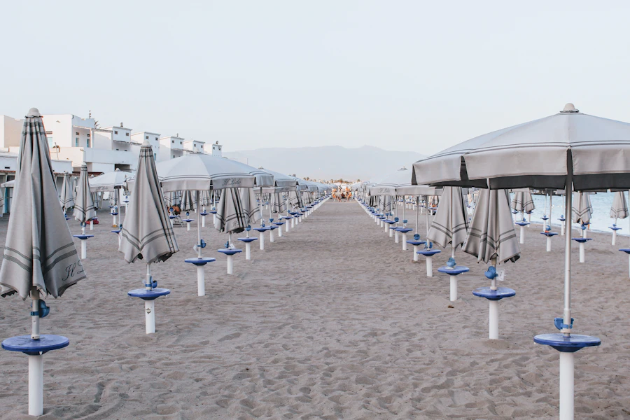 Neatly aligned closed umbrellas and lounge stands stretch down a sandy beach walkway toward the horizon, with resort buildings on the left and calm water on the right. Soft light and hazy mountains create a quiet, orderly seaside scene.