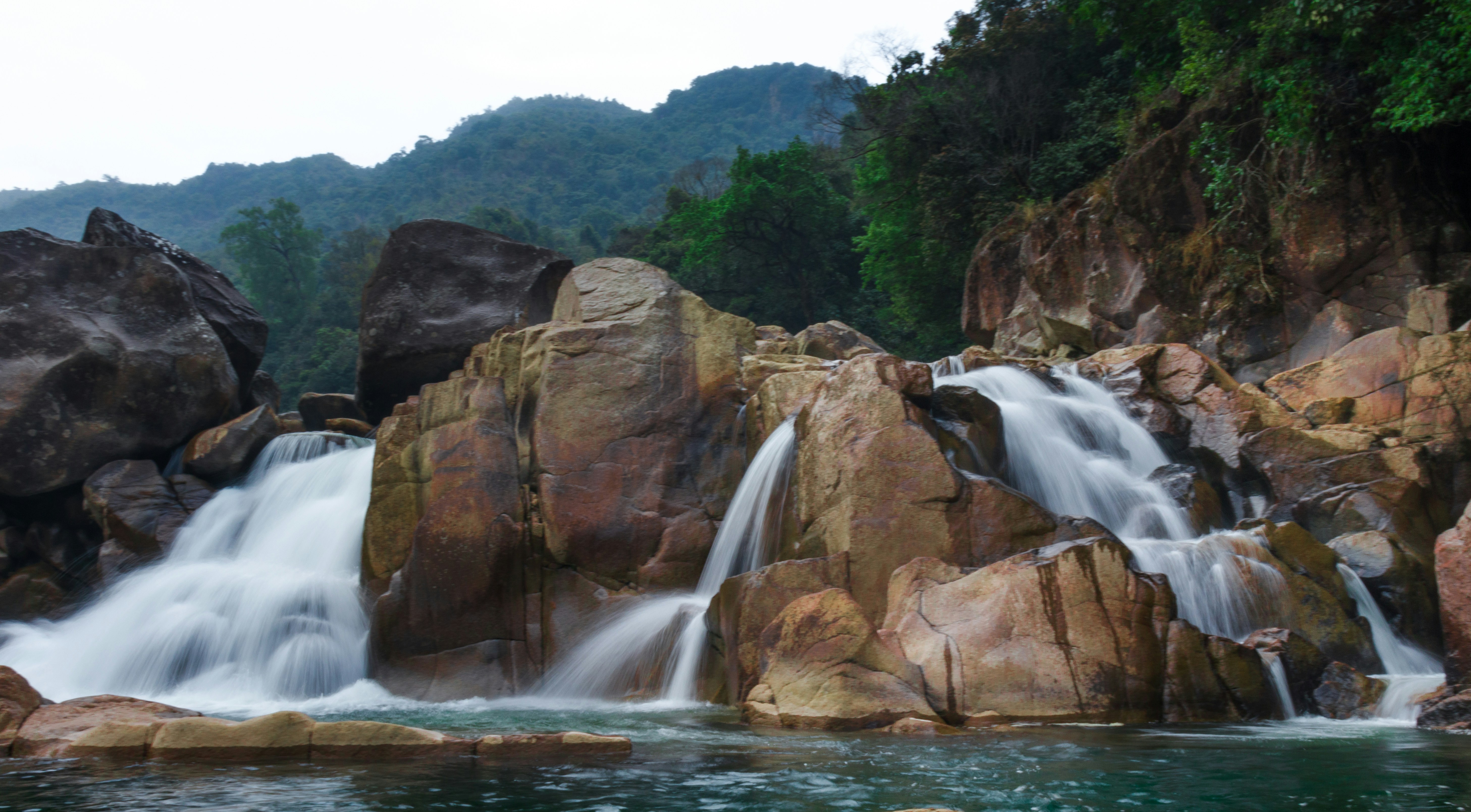 Gentle waterfalls cascading over rugged rocks, surrounded by lush greenery and distant mountains.