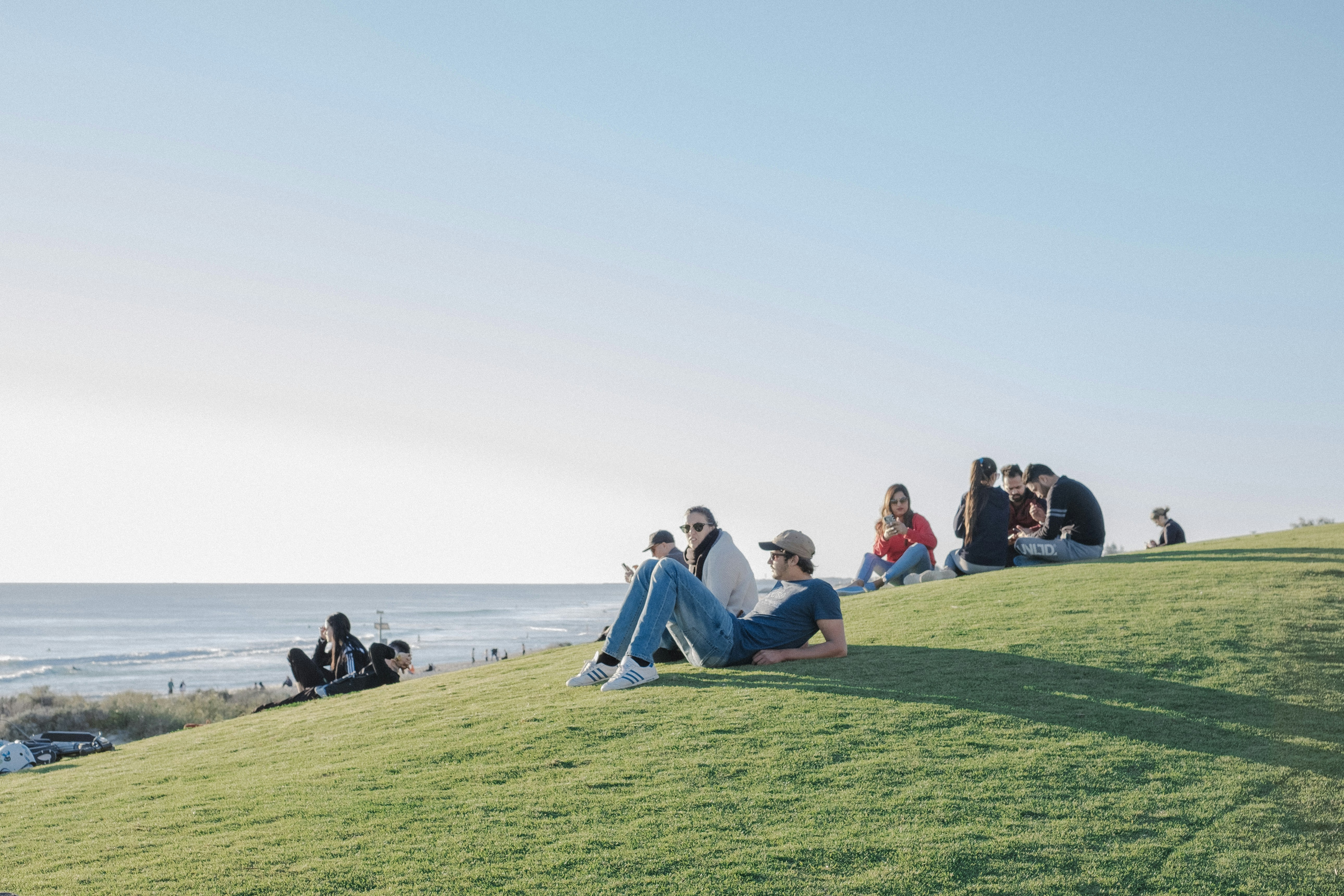 People relaxing on a grassy hill overlooking the ocean under a clear blue sky.