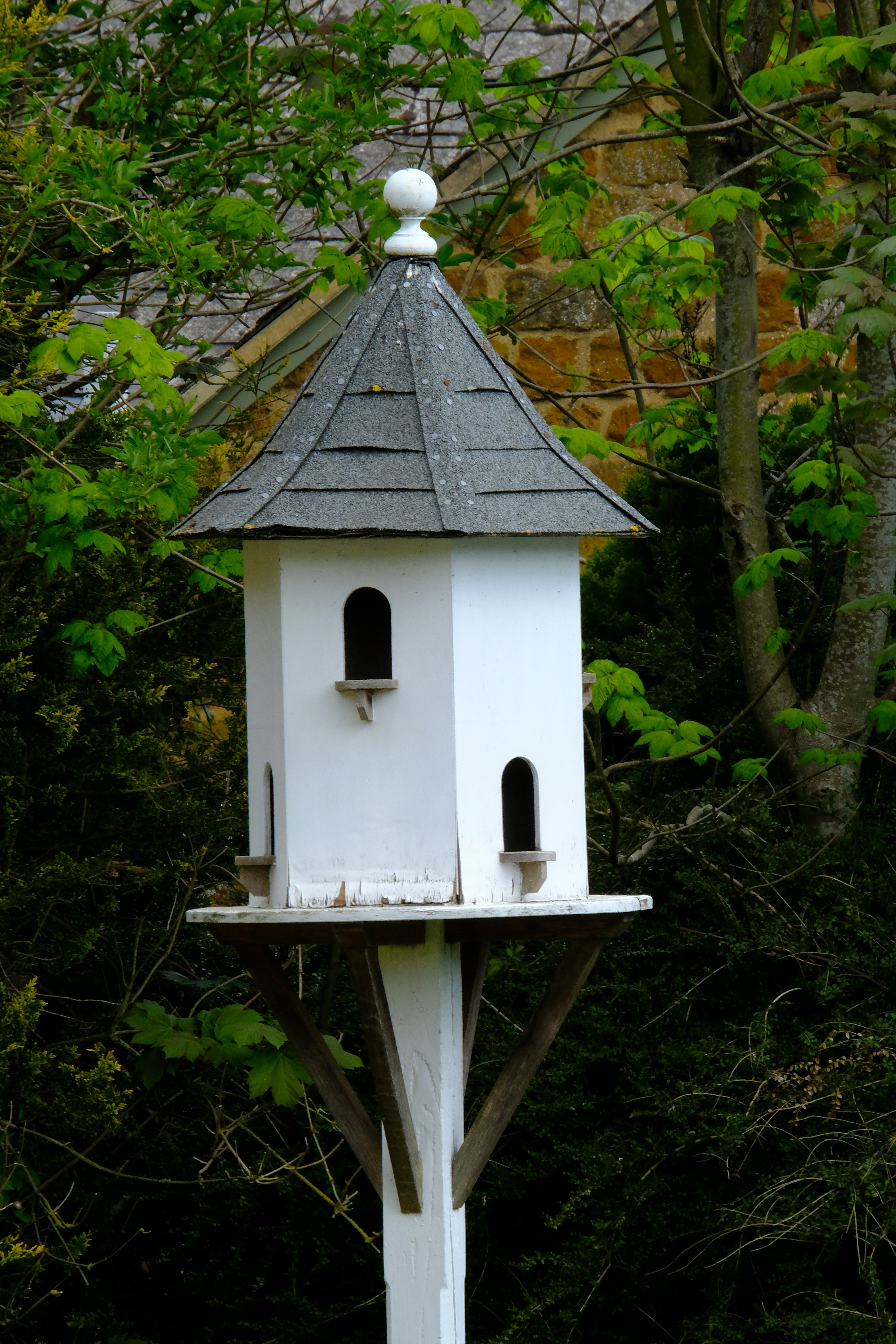 Elegant white birdhouse perched on a wooden post, surrounded by vibrant green leaves and rustic stone background.