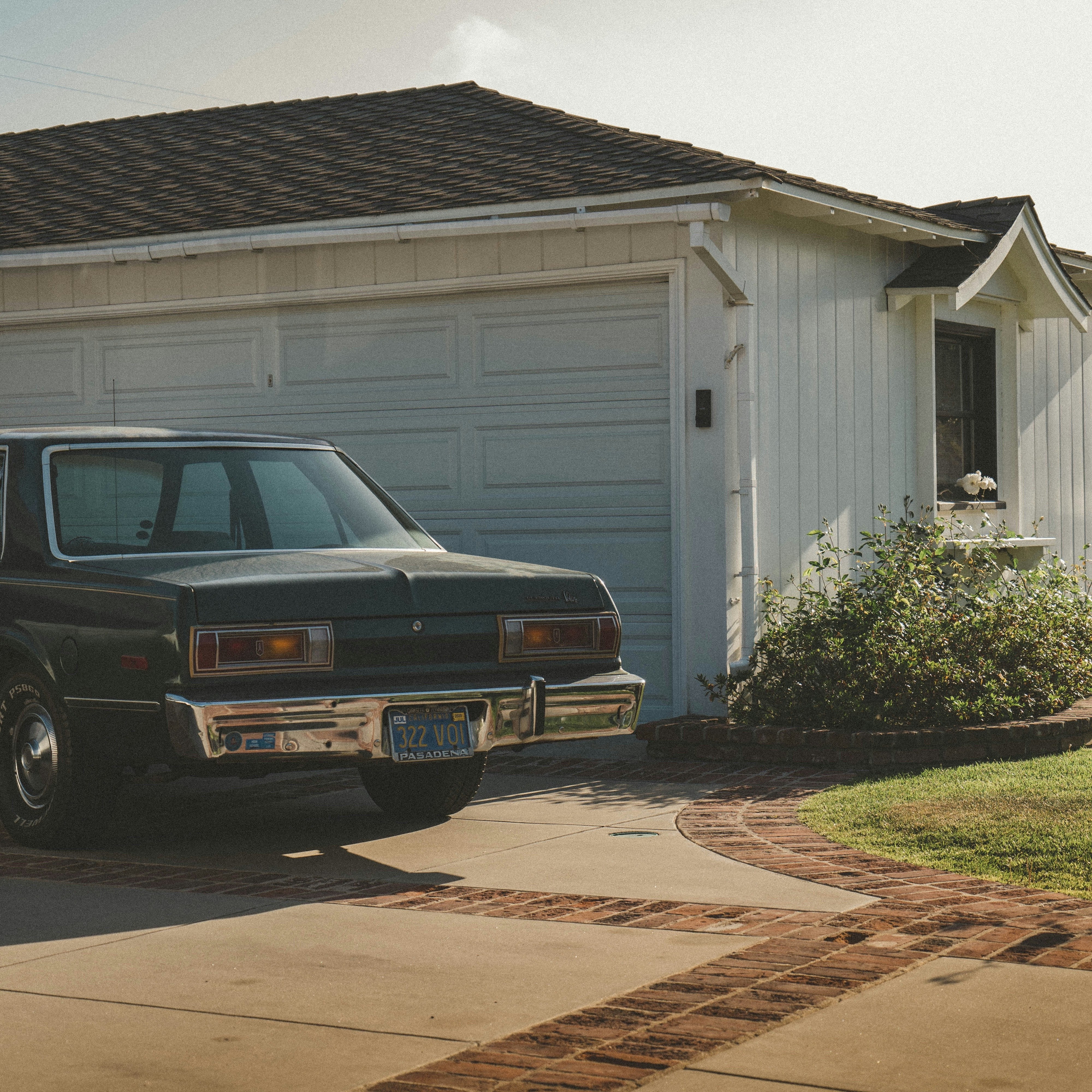 Classic green sedan parked in a sunlit driveway, framed by a charming home and lush greenery.