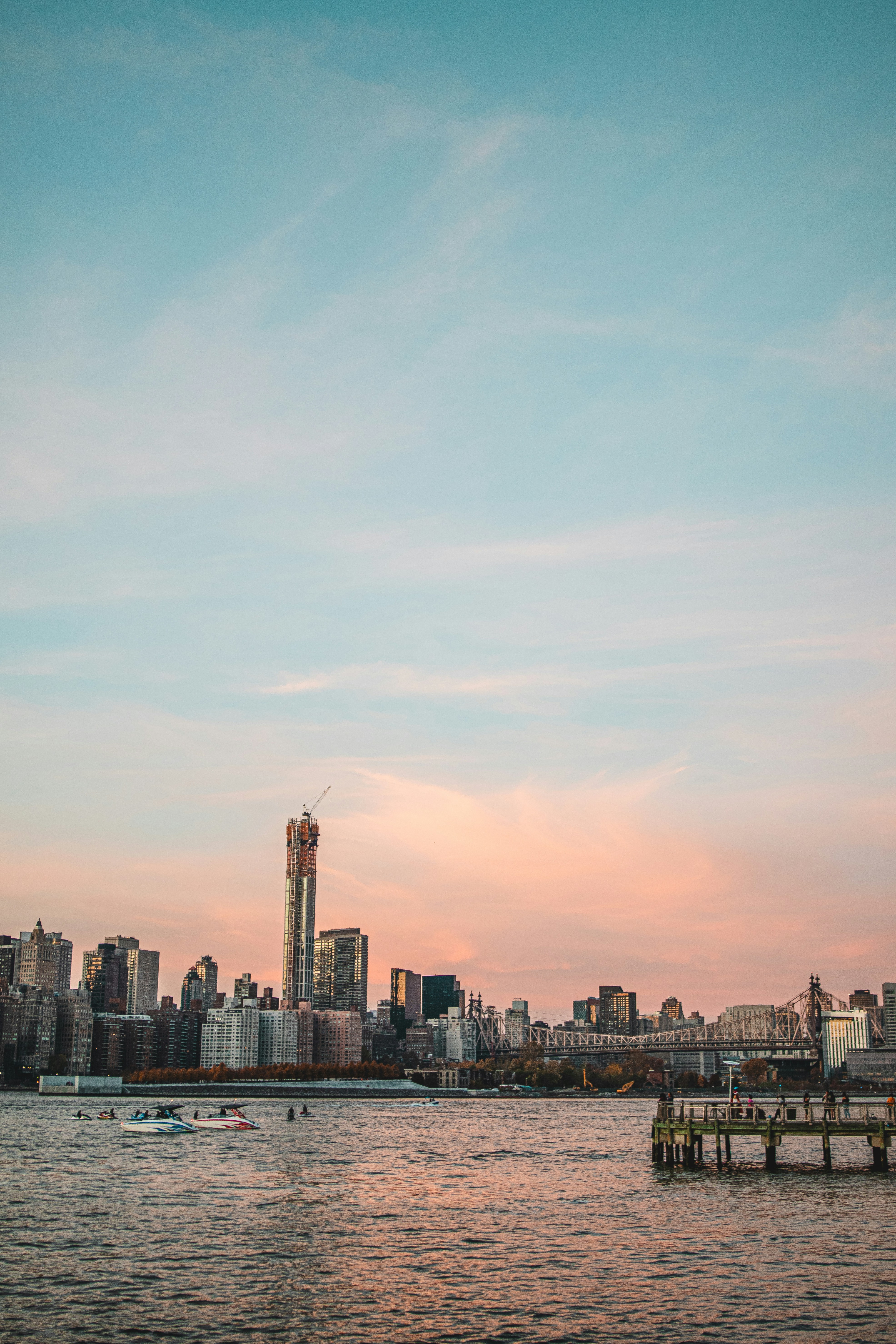 City skyline at dusk with a prominent skyscraper under construction and boats on the water. Soft pastel colors in the sky enhance the urban landscape.