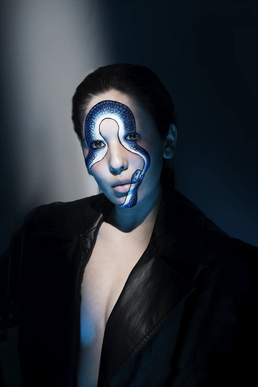 Woman in dramatic low-key portrait lighting with a blue snake-like face painting and dark clothing. Her face is centered against a shadowy background.