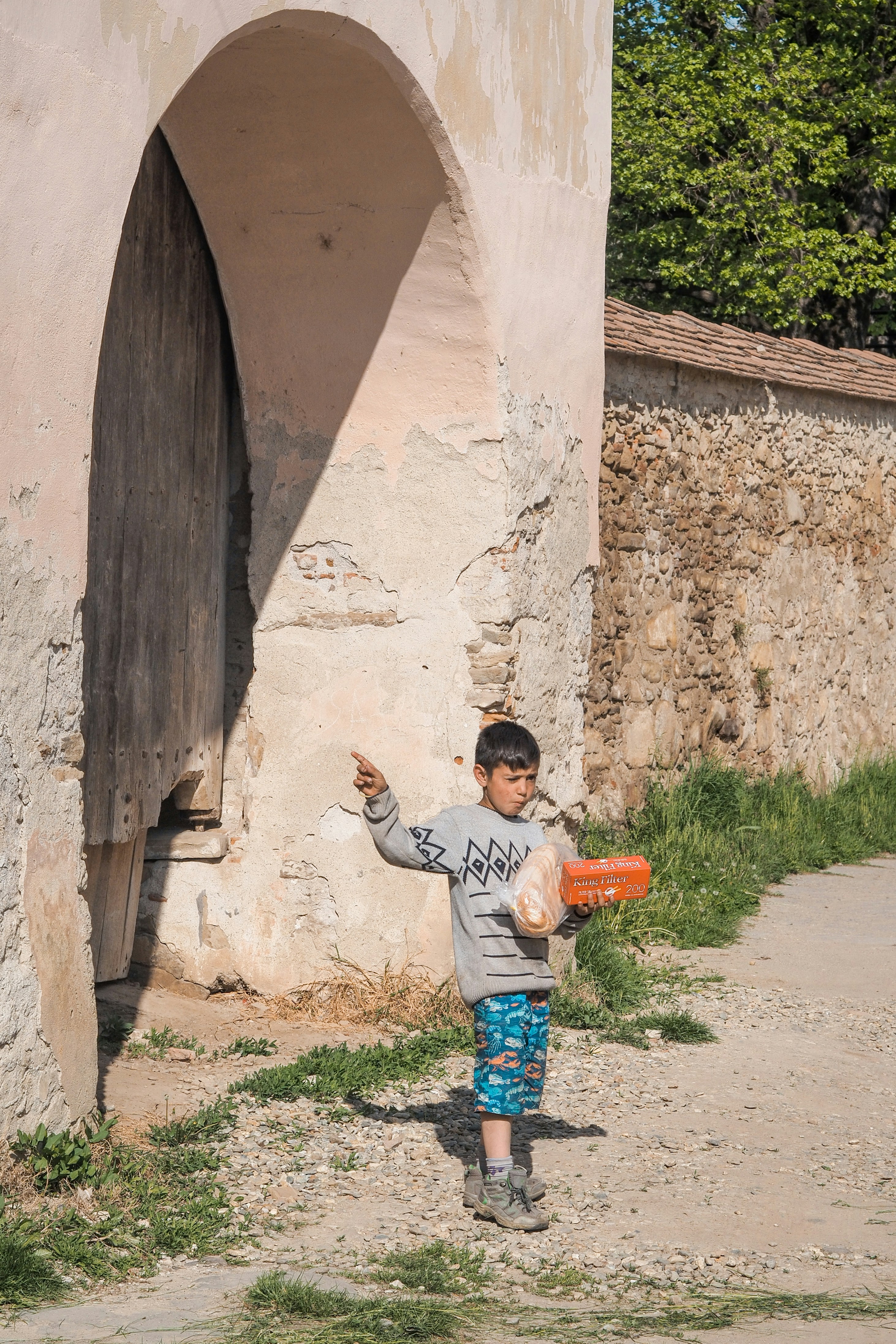 Sunlit Archway Moment: A Boy Points with an Orange Box by a Weathered Wall