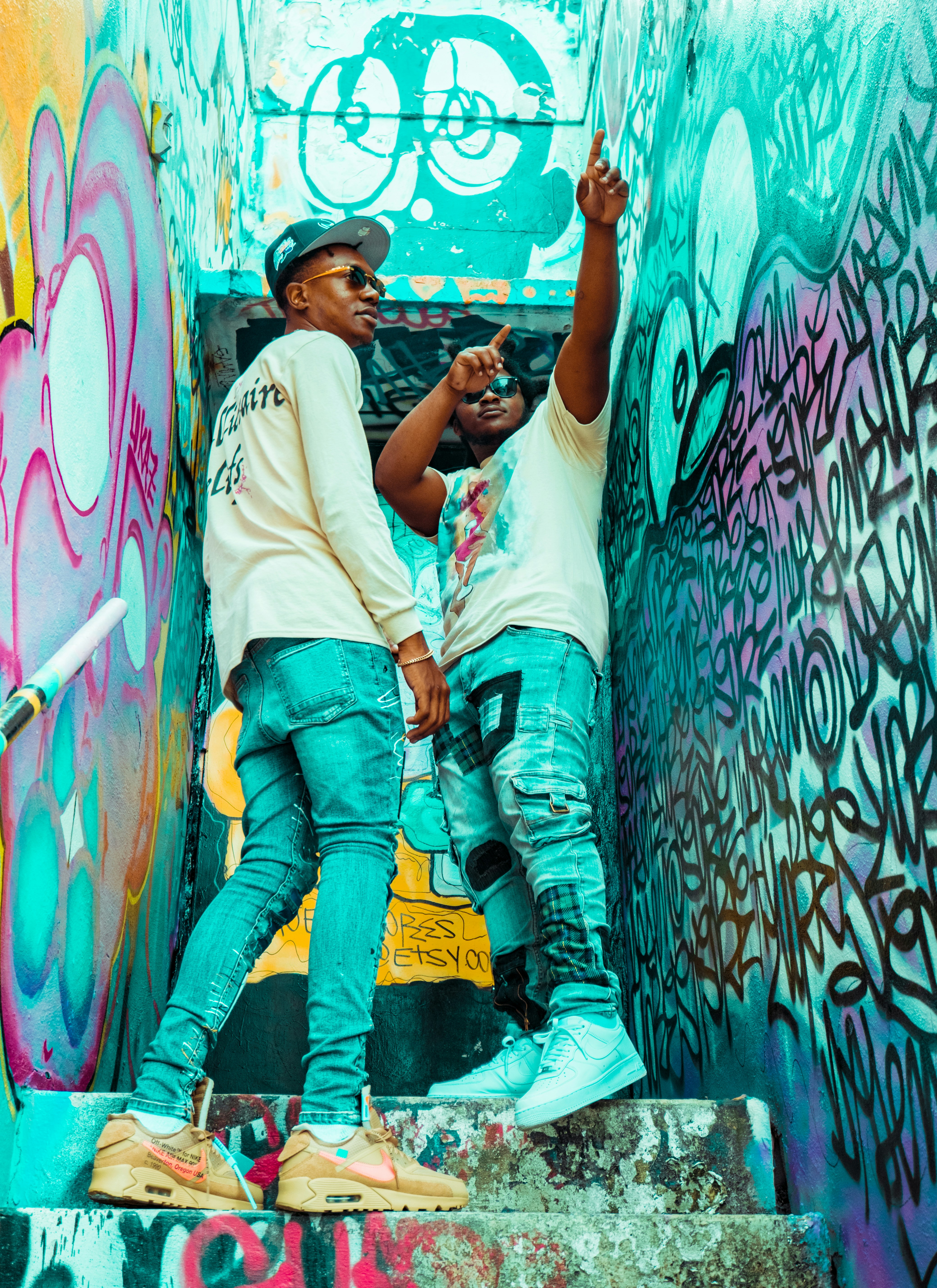 Two individuals pose confidently in a vibrant graffiti-filled stairwell, showcasing urban fashion and expressive gestures.