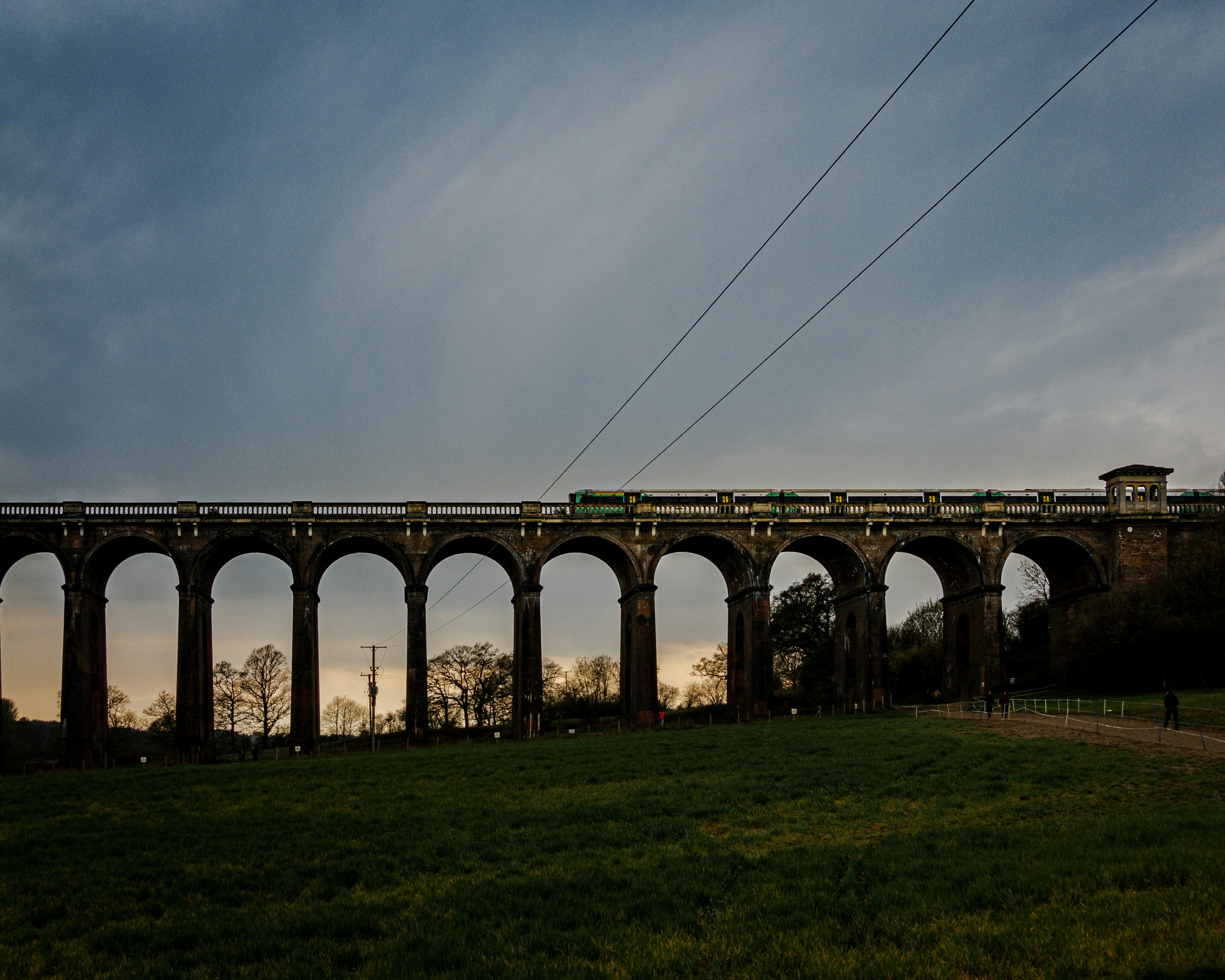 A historic railway viaduct stretches across the landscape, with a train traversing its arches under a moody sky.