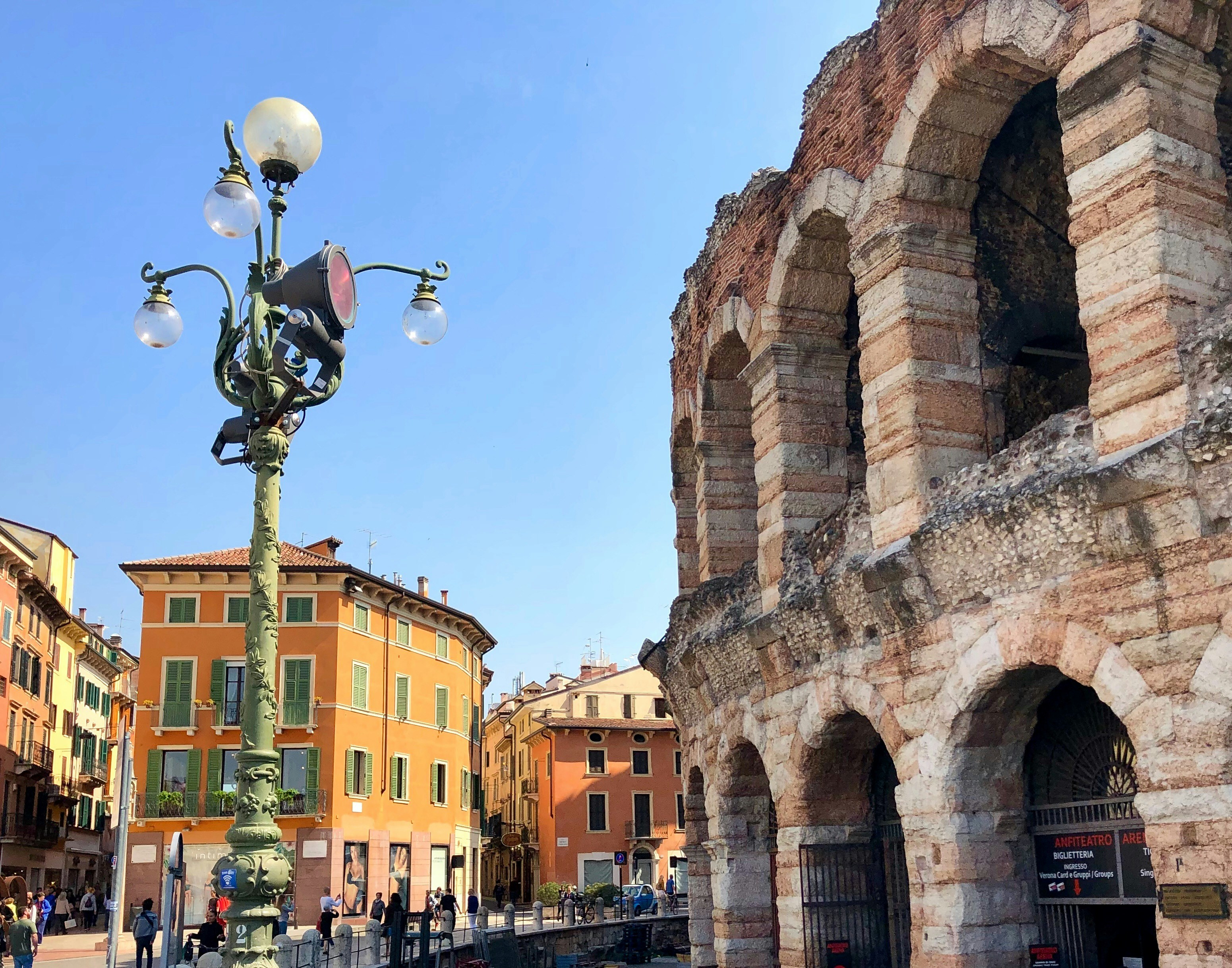 Ancient stone amphitheater with tall arches beside colorful historic buildings under a clear blue sky.