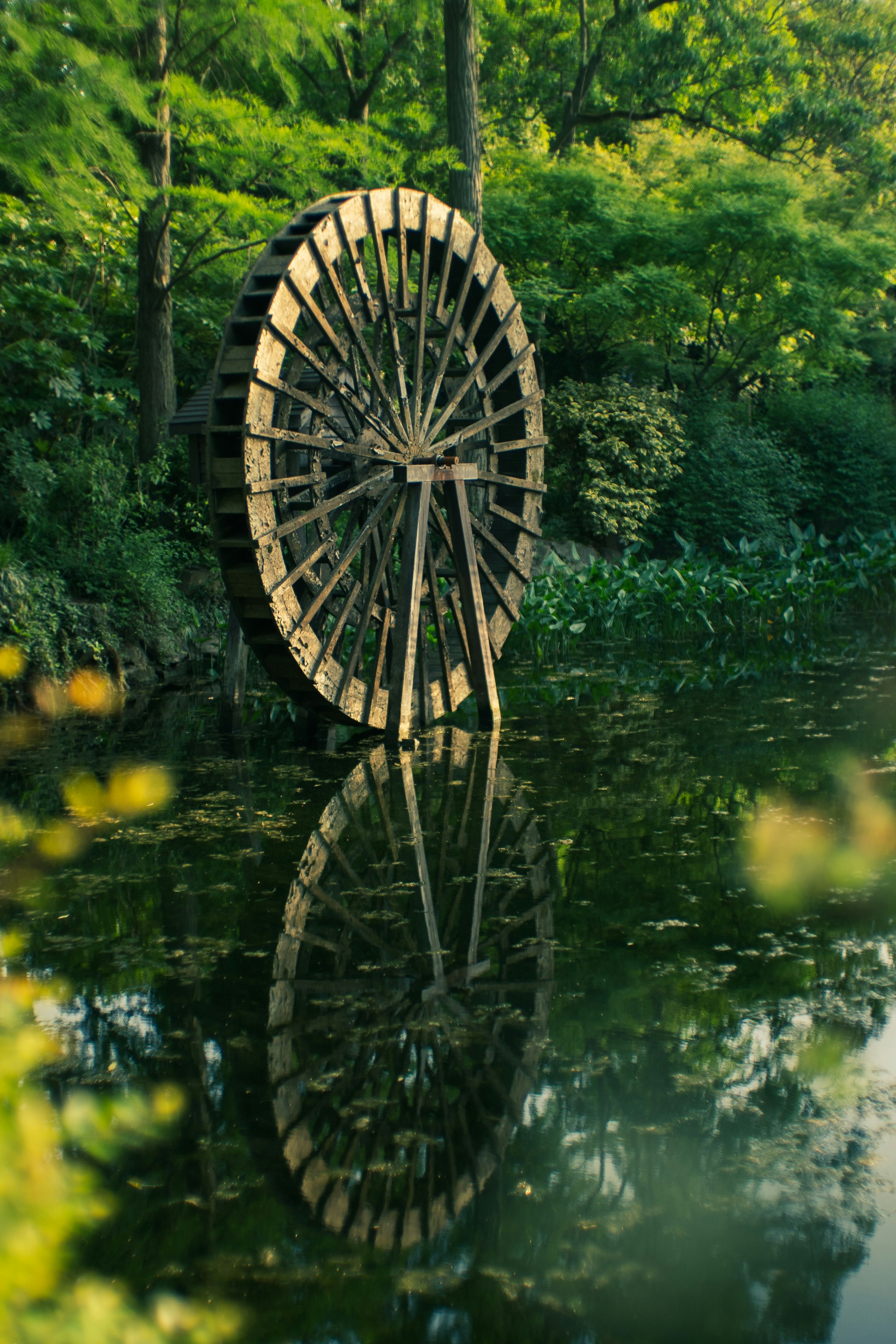 A rustic water wheel stands beside a tranquil pond, its reflection shimmering in the still water surrounded by lush greenery.