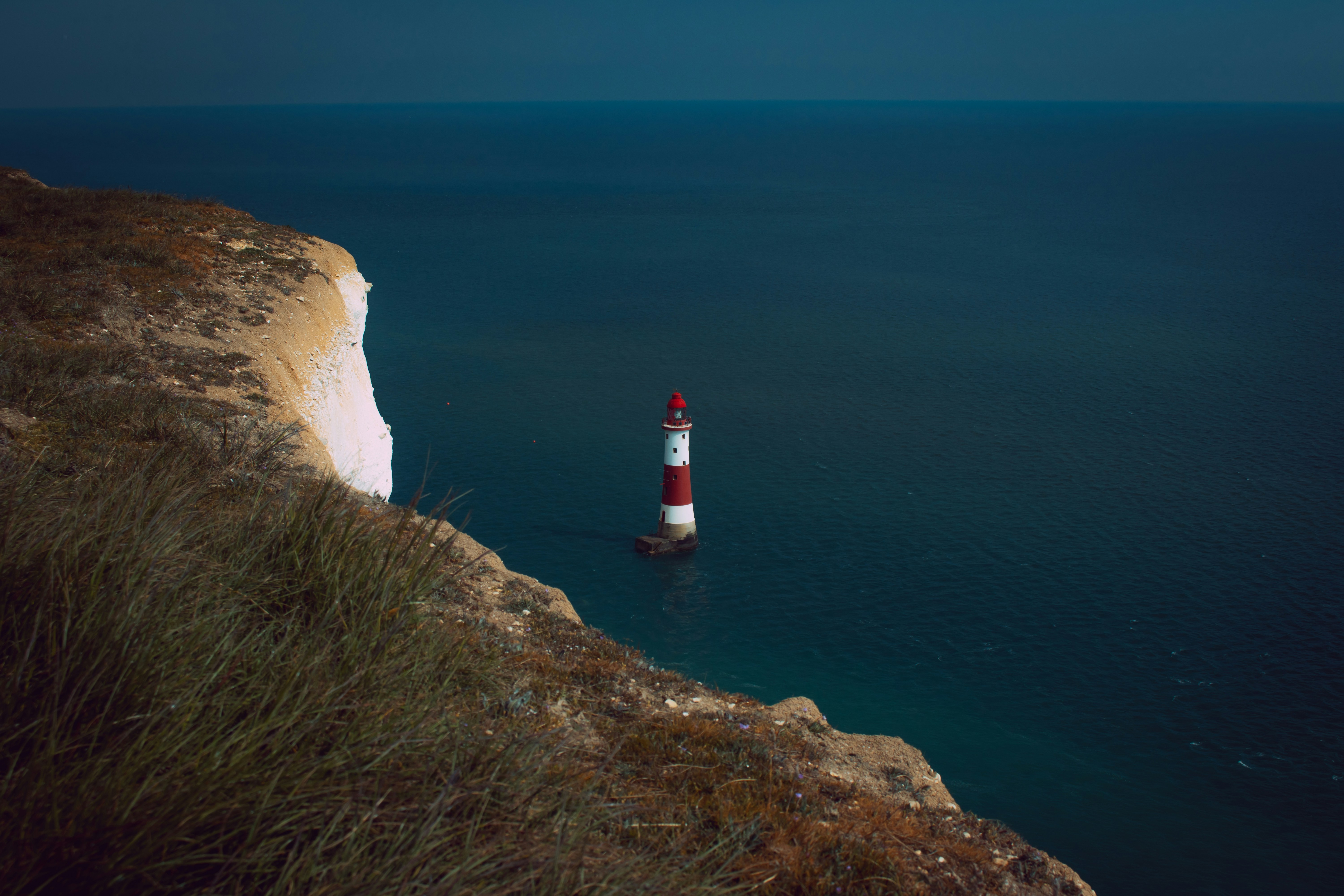 Lighthouse standing amidst calm blue waters at dusk, viewed from a grassy cliff edge.