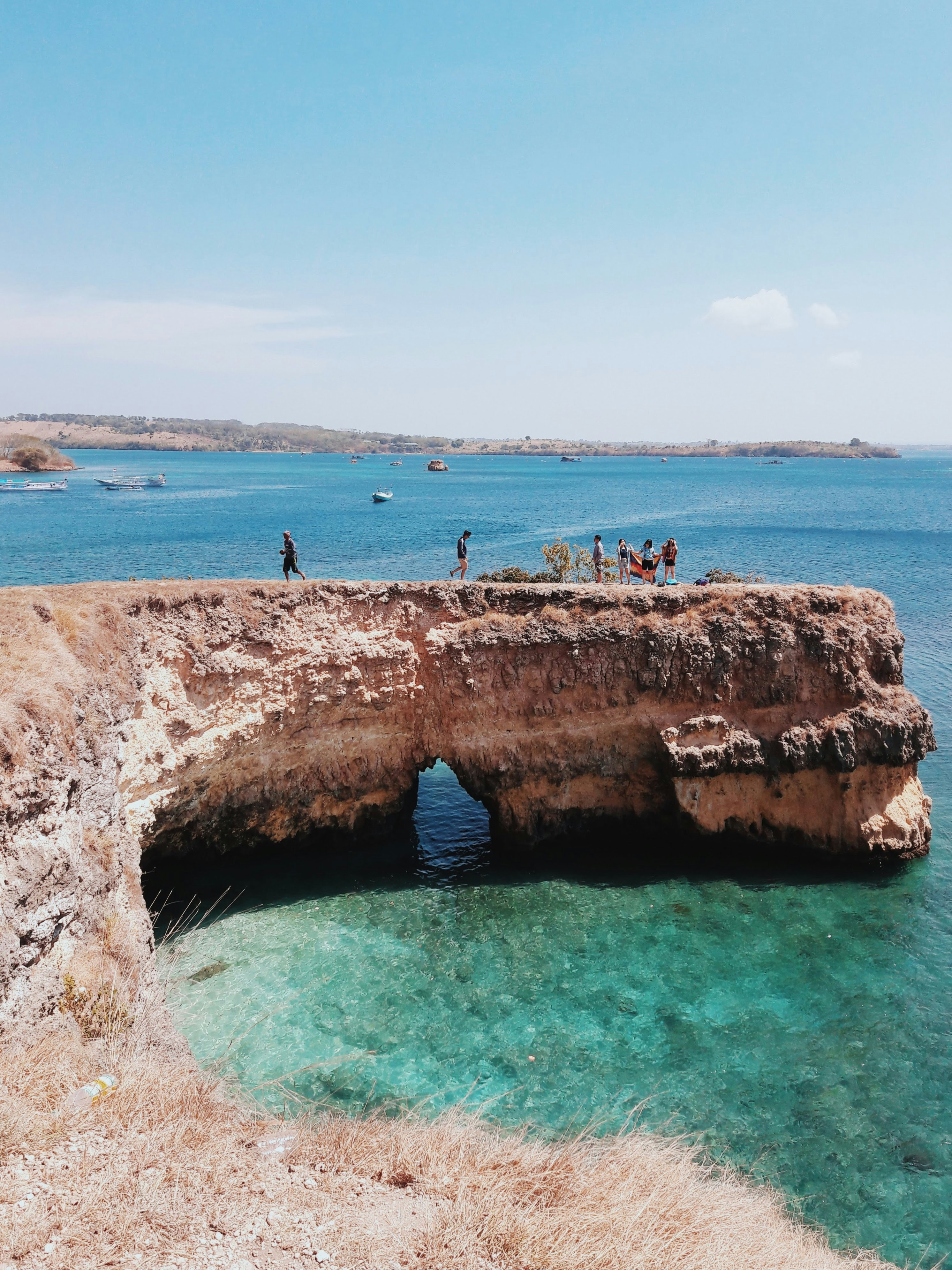 Sunlit coastal scene showing a weathered sea arch extending over clear turquoise water with a rugged shoreline. People stand along the arch’s edge, taking in the view.