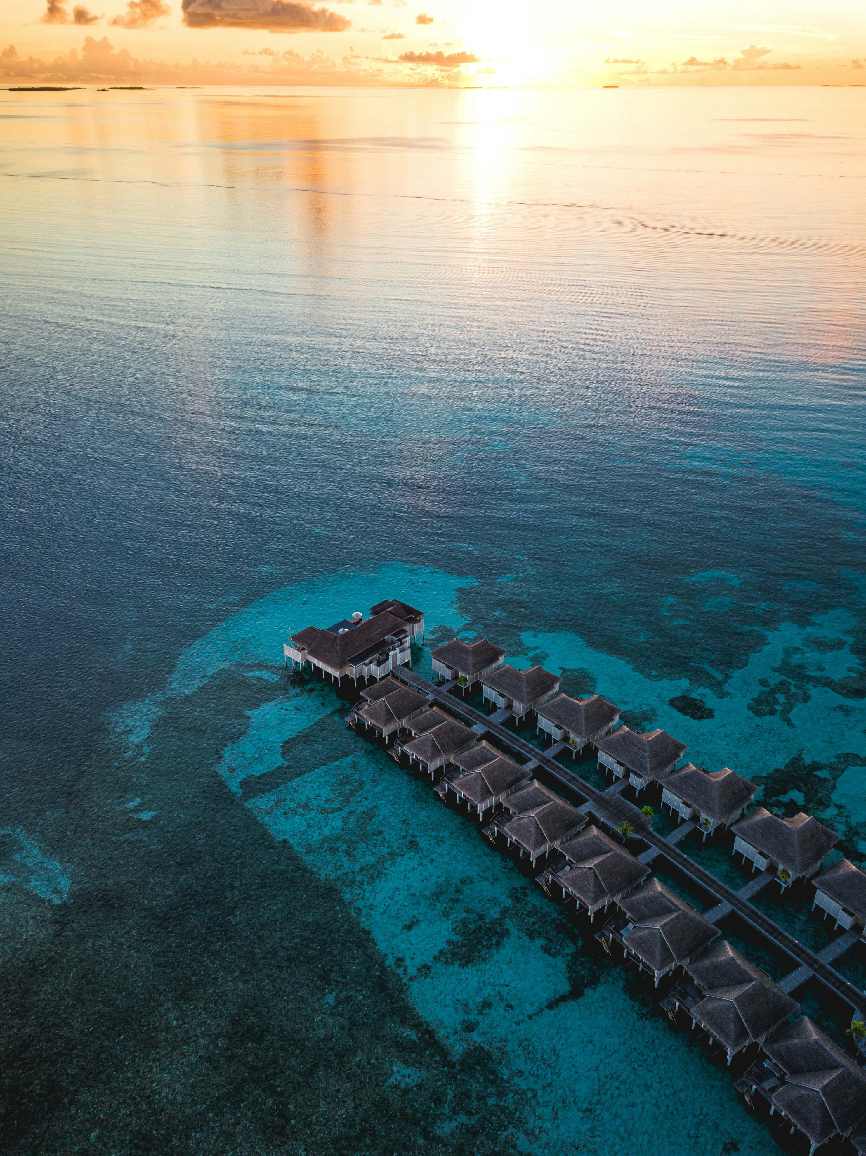 Aerial view of luxurious overwater bungalows nestled in a vibrant coral lagoon at sunset, reflecting warm hues on the tranquil water's surface.