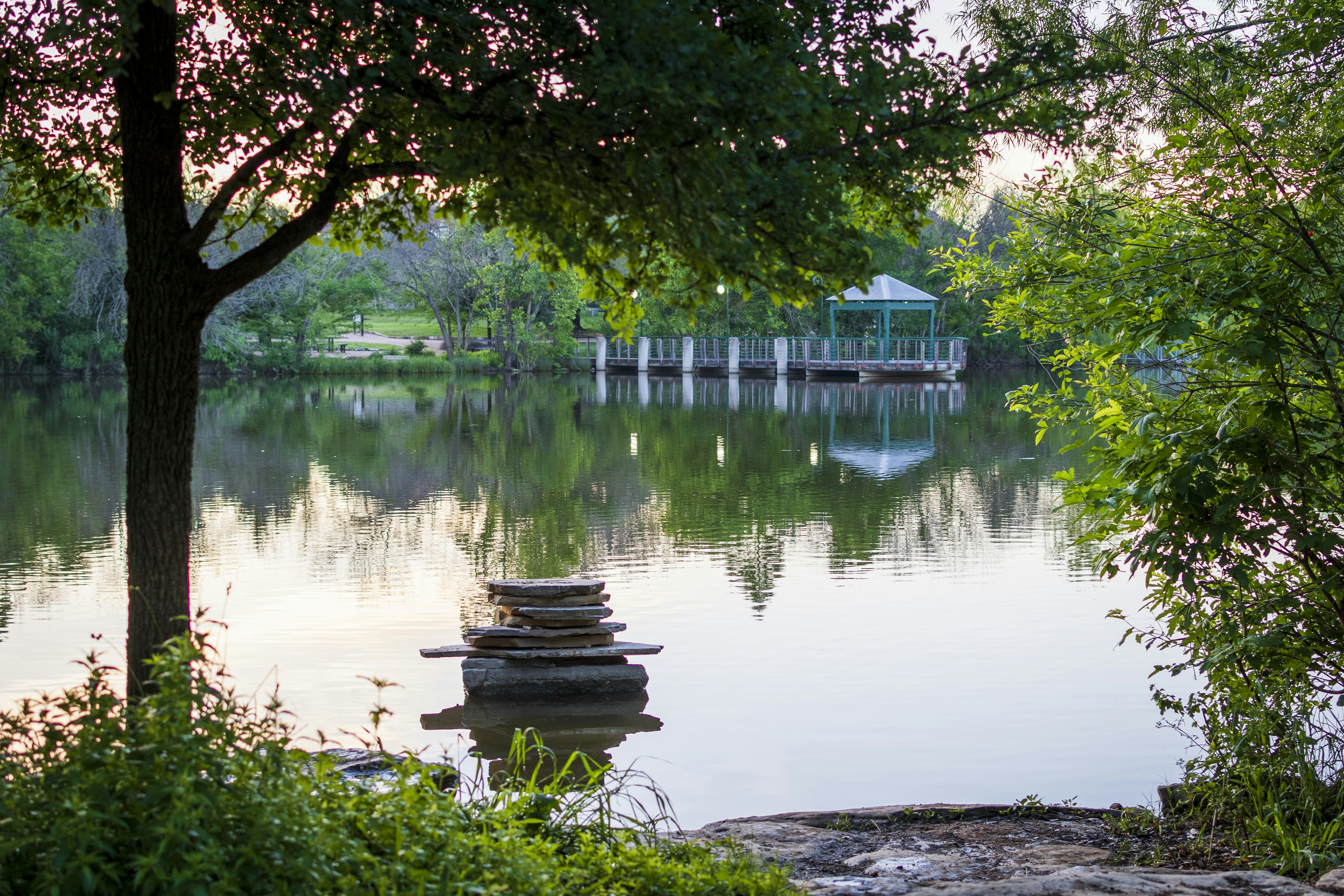Still lake reflecting a gazebo and surrounding greenery, framed by trees and rocks in the foreground.