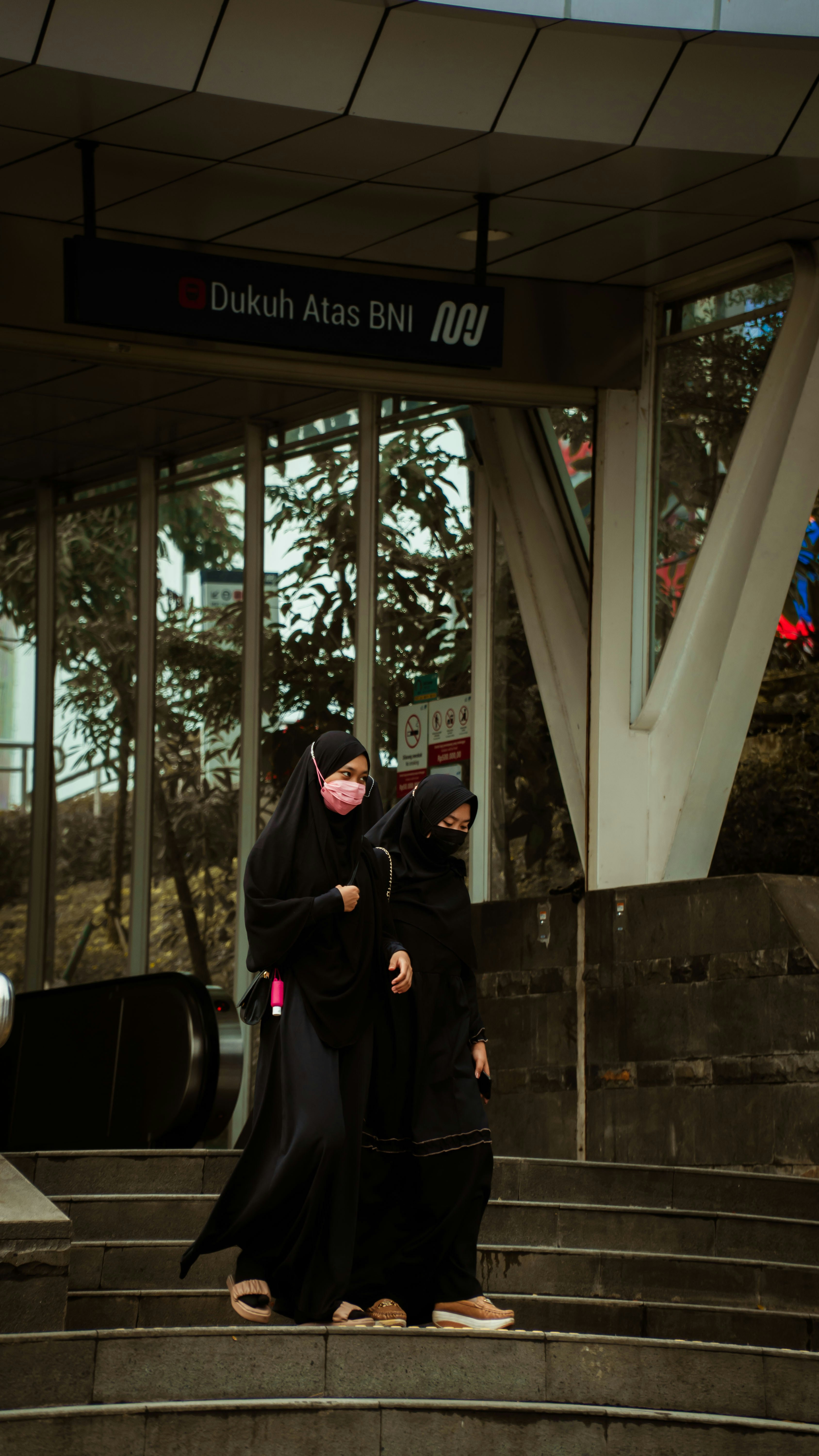 Two women in traditional attire descend a staircase near a transit station, embodying the blend of cultural heritage and urban life.