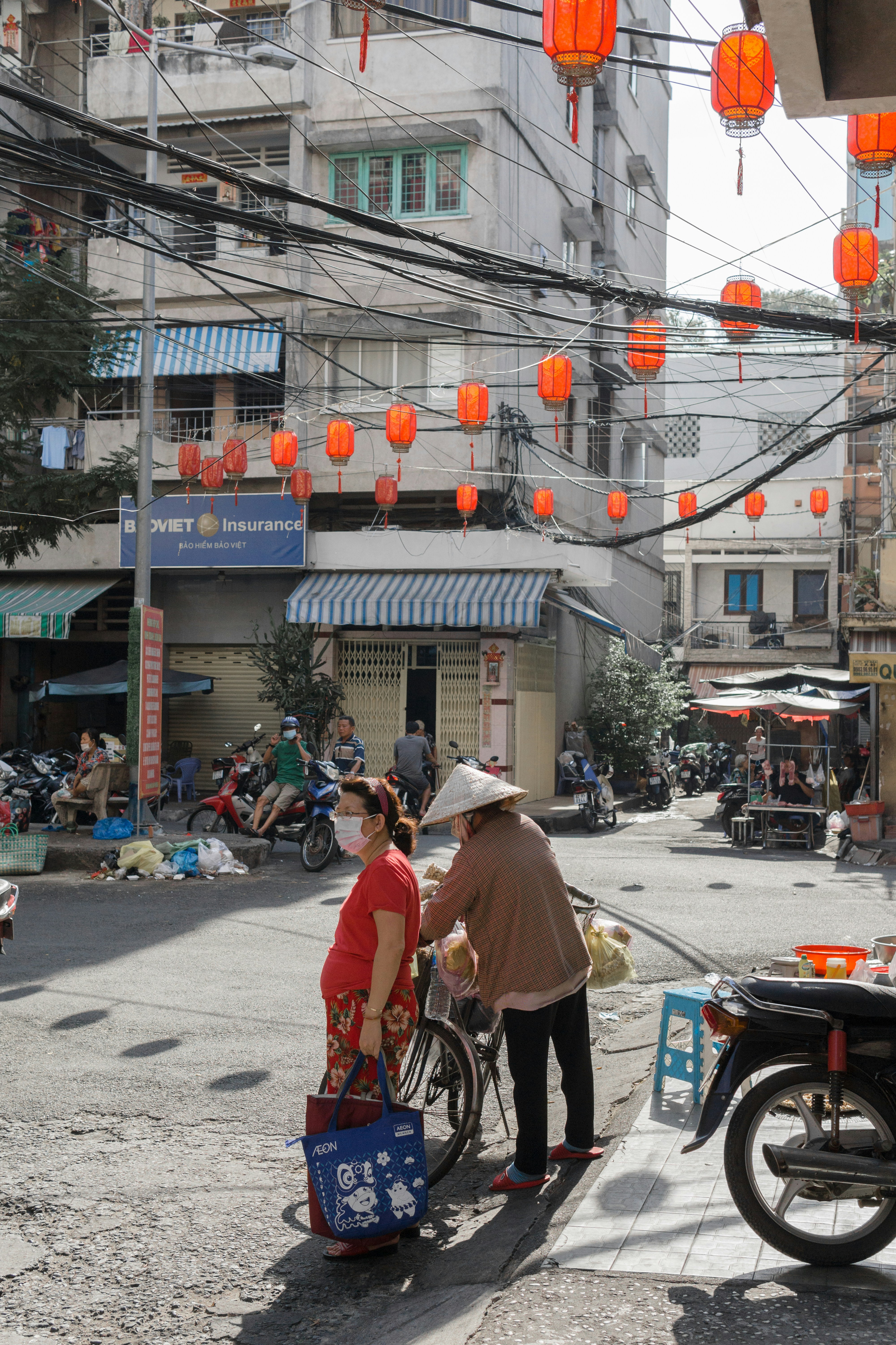Two women stand by a bicycle, surrounded by vibrant red lanterns and bustling street activity. The scene captures the essence of local culture and daily routines.