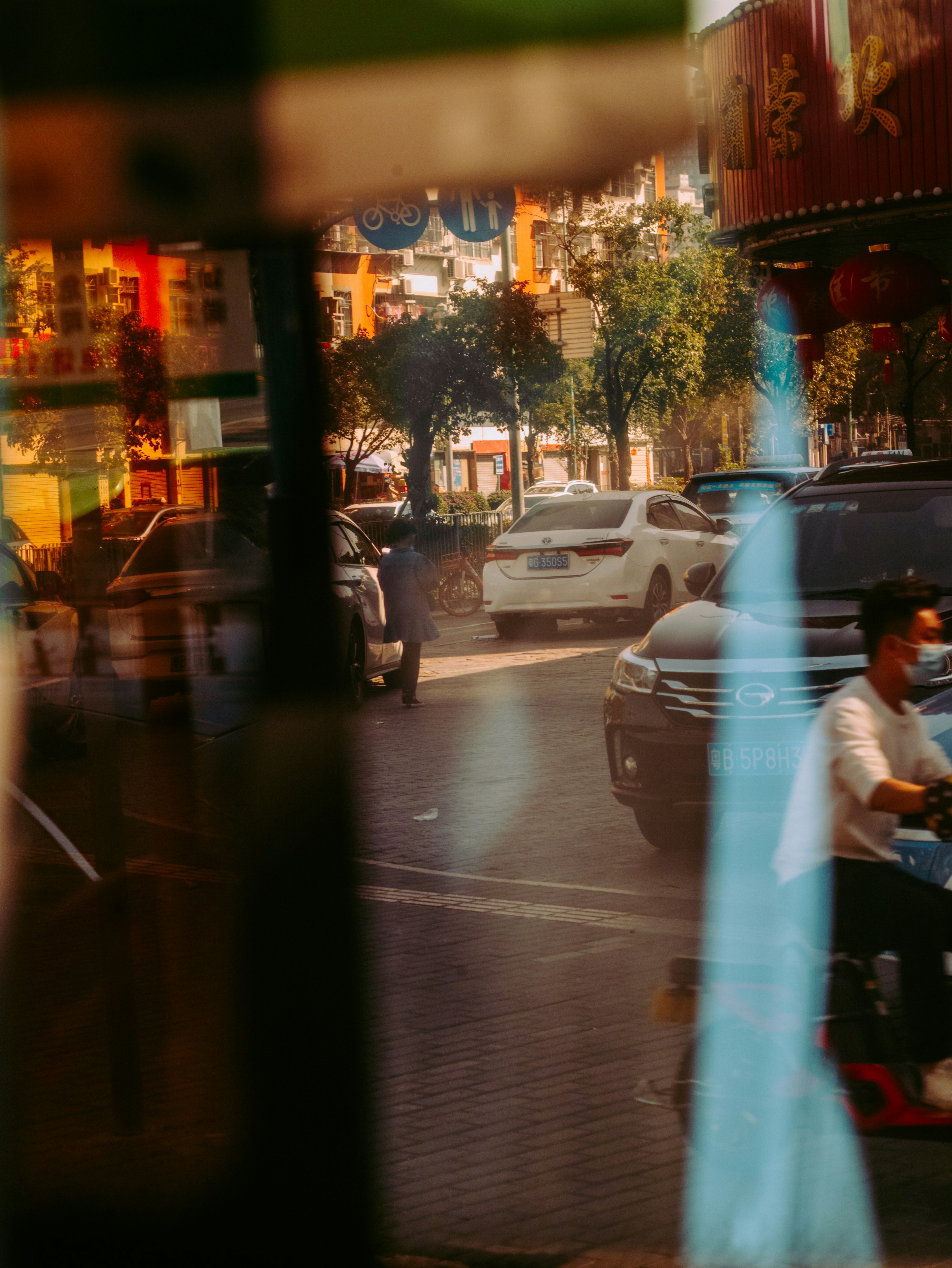 A busy city street viewed through a reflective storefront window, creating layered reflections and depth. A white car and pedestrians occupy the mid-ground under warm afternoon light.