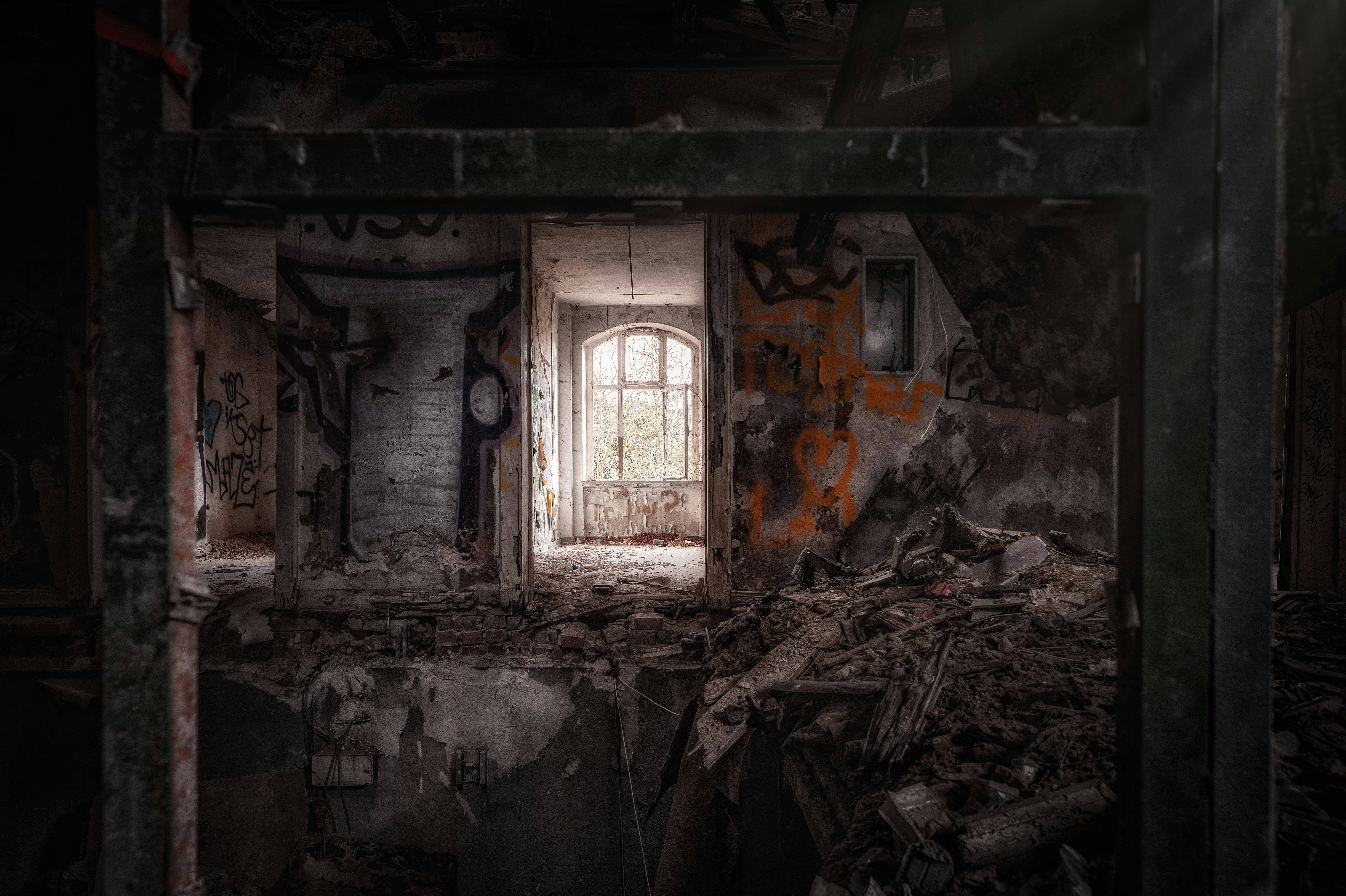 An interior view of a dilapidated room, framed by a doorway, revealing a sunlit window amidst scattered debris and graffiti.
