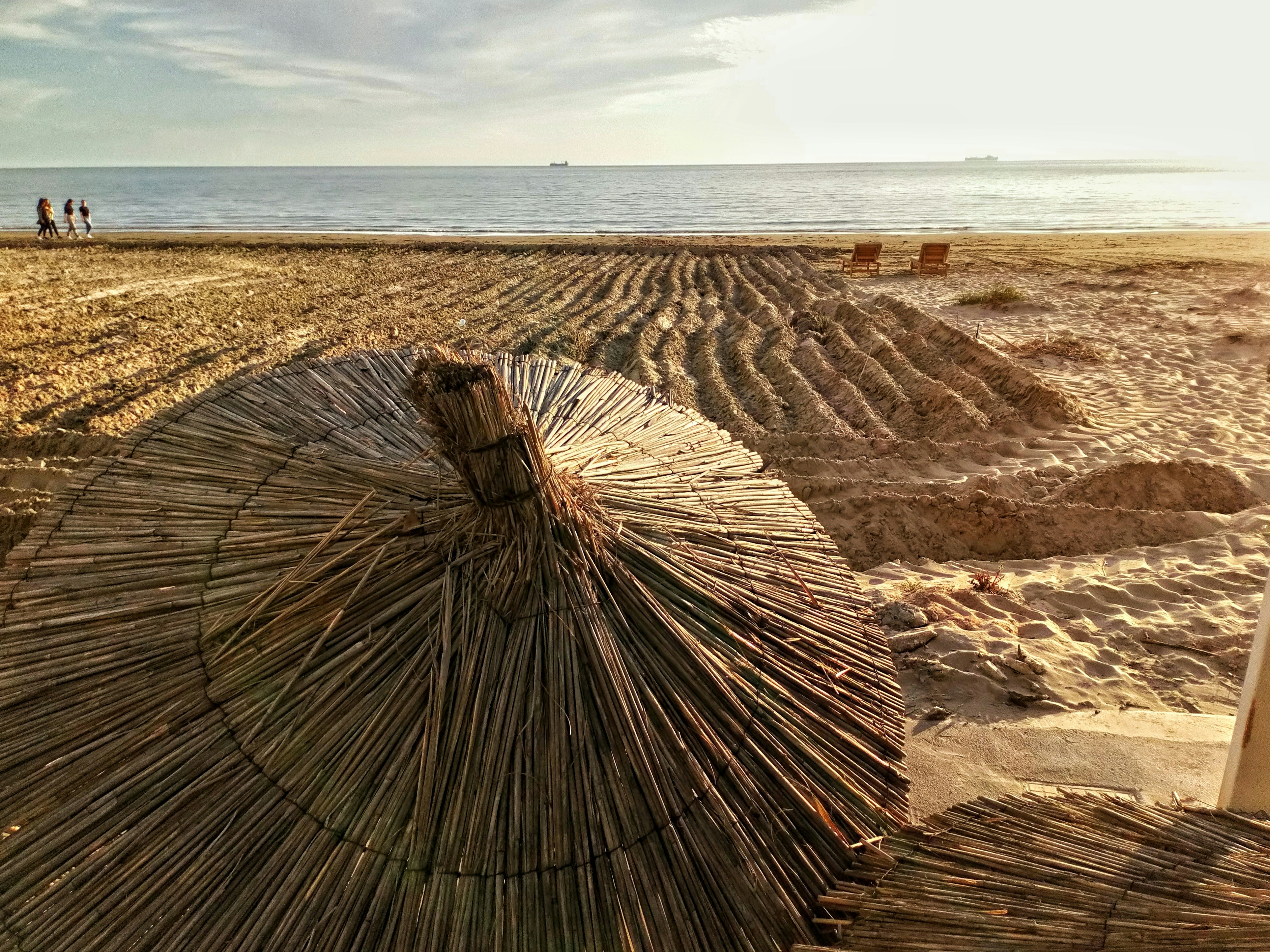 Sunlit beach scene with a large thatched umbrella in the foreground and sand ripples leading to a calm sea on the horizon.