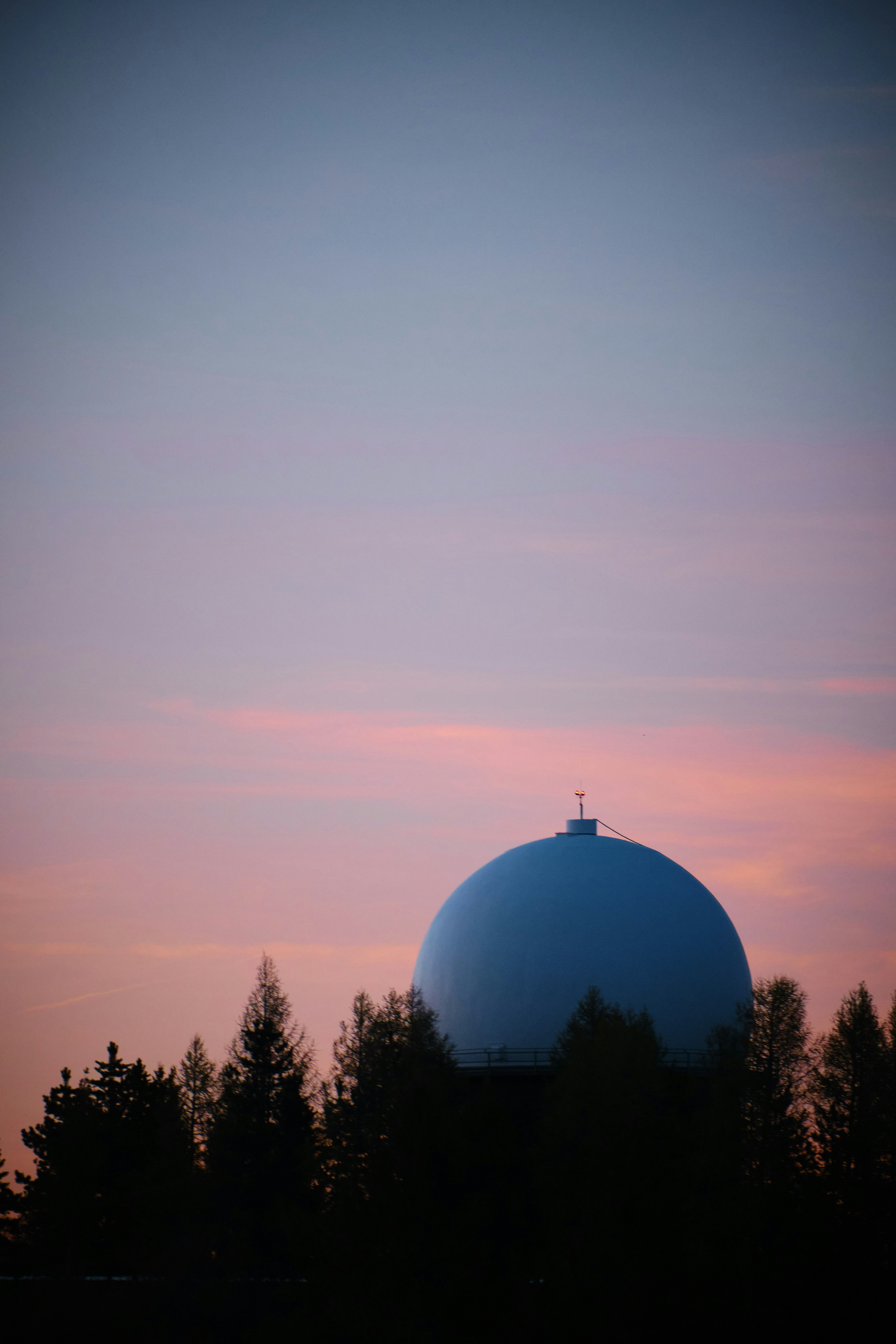 A large dome structure silhouetted against a pastel sky at twilight, framed by dark treetops. The scene evokes a sense of tranquility and mystery.