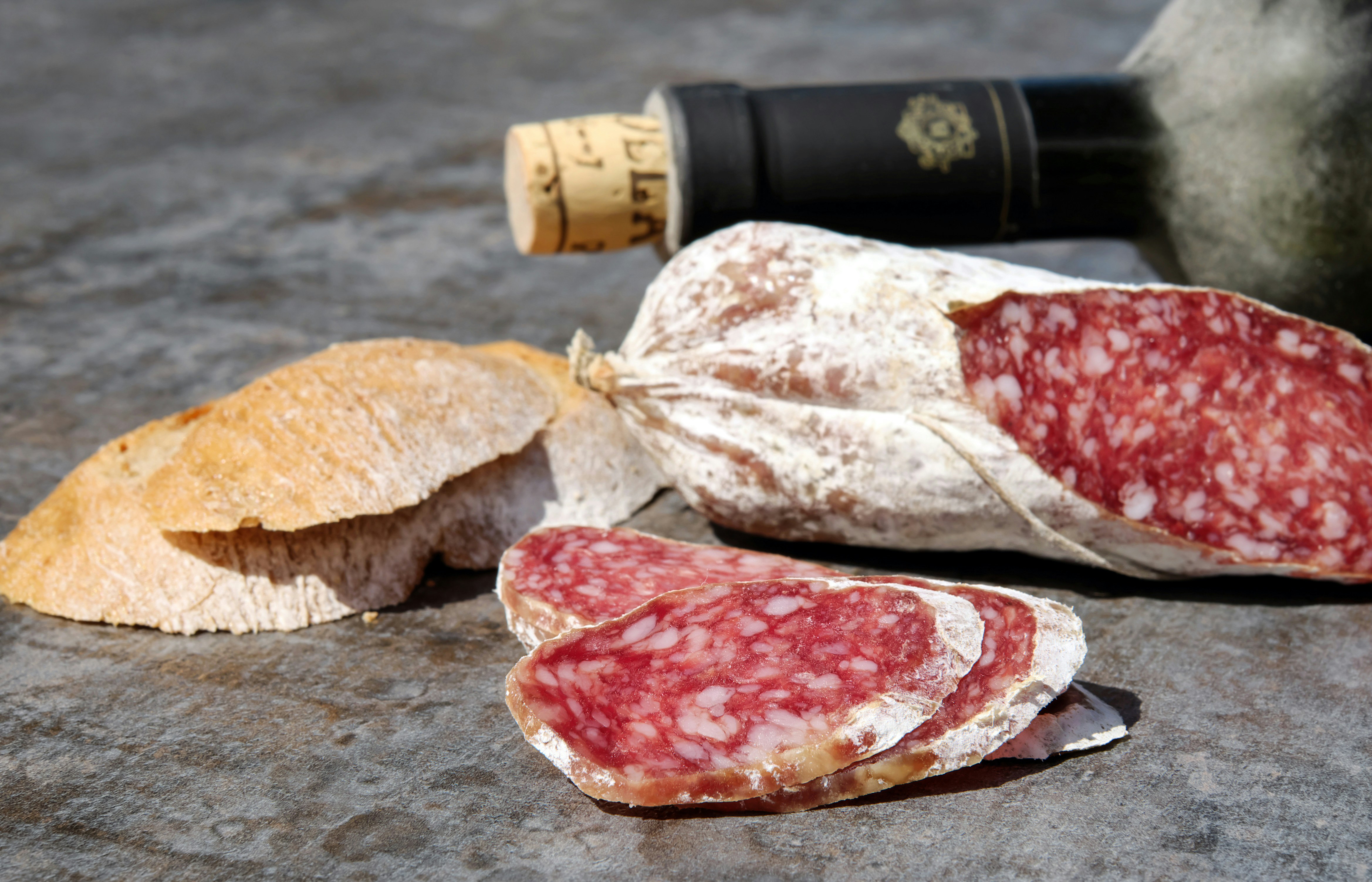 Slices of cured meat and bread on a stone surface with a wine bottle in the background.