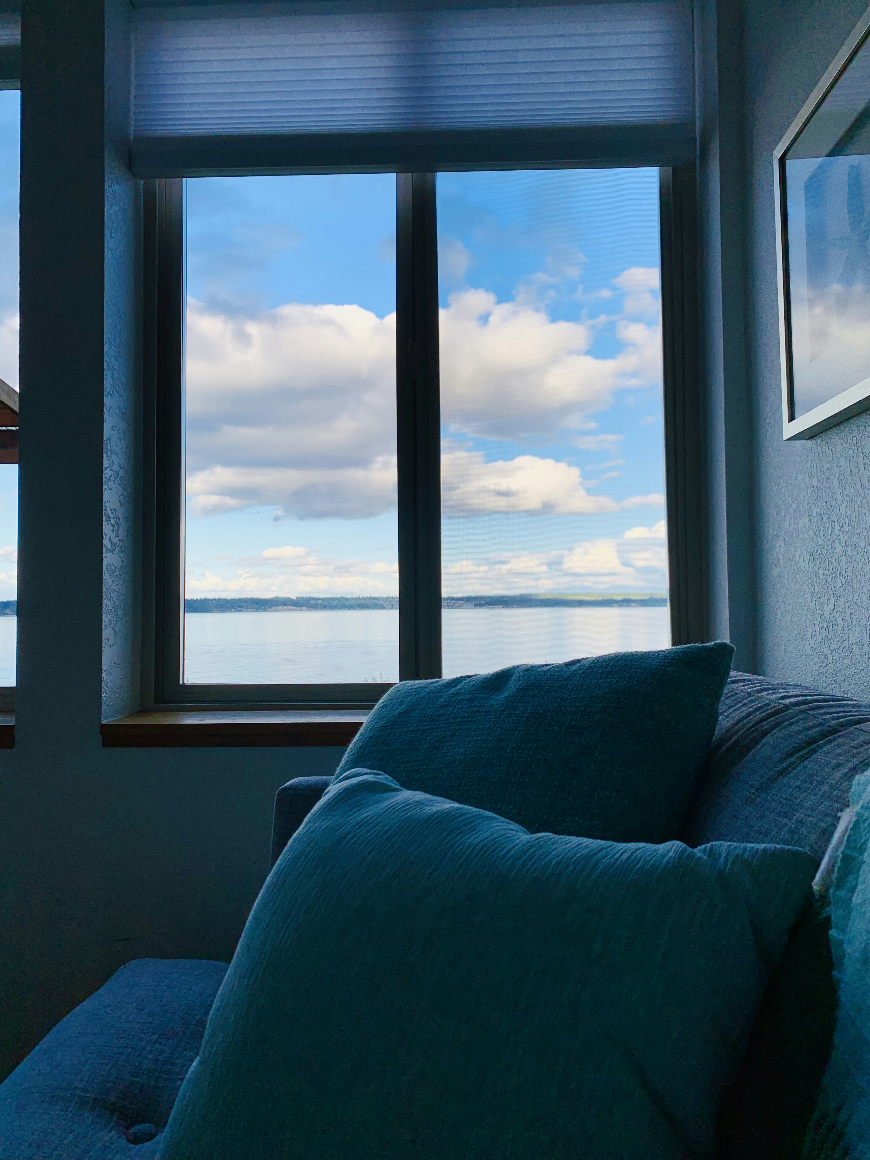 A serene view of calm waters and fluffy clouds through large windows, complemented by a plush couch adorned with soft pillows.