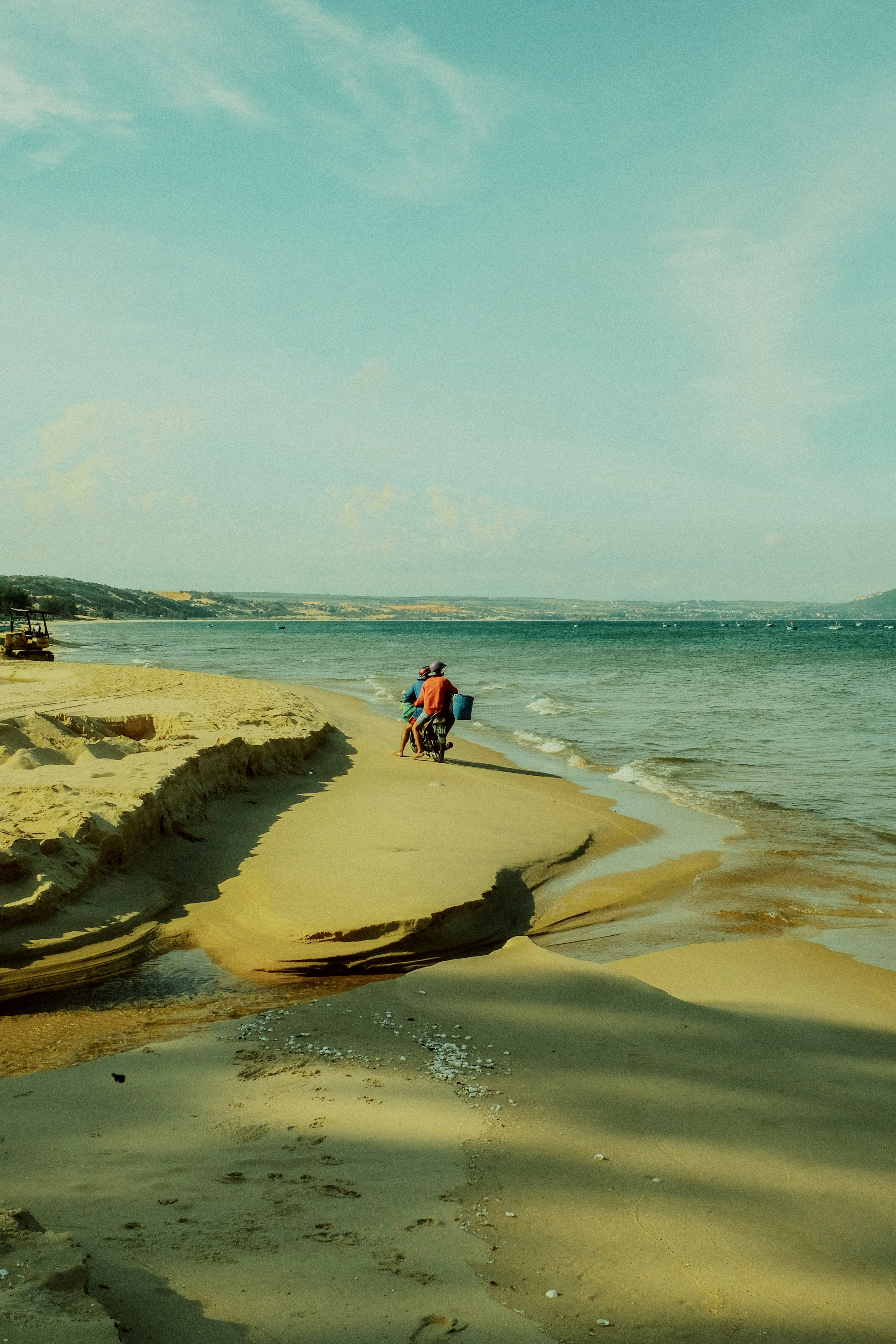 Two individuals leisurely walking along a sandy beach, with gentle waves lapping at the shore under a clear sky.