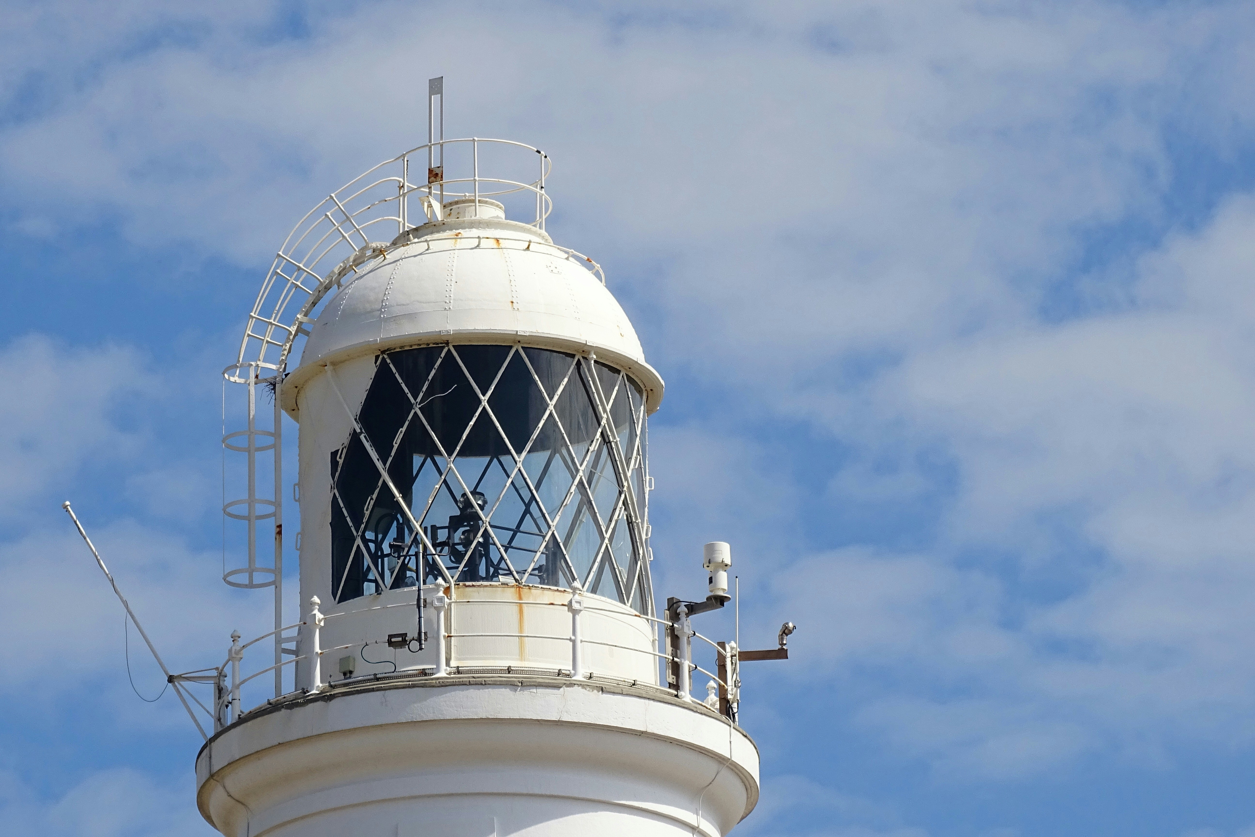 Lighthouse lantern room featuring intricate glass panes and metalwork, set against a backdrop of blue sky with scattered clouds.