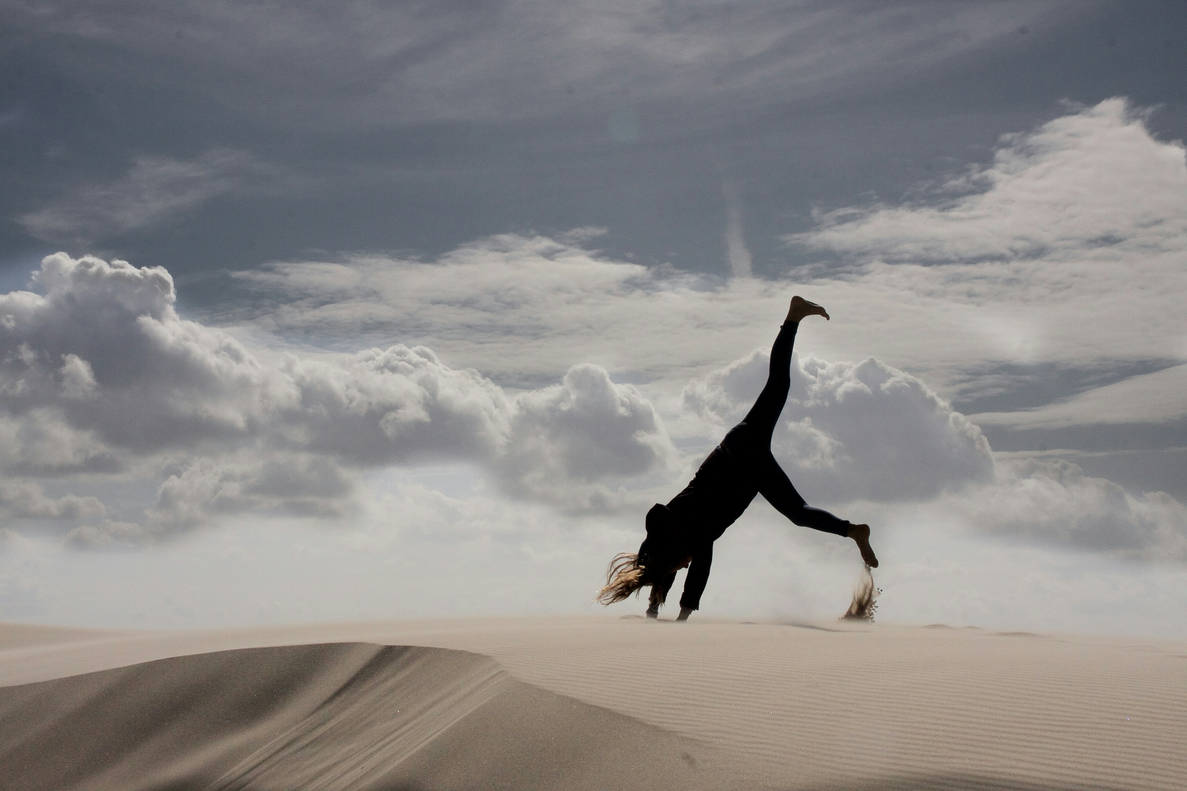 Silhouette of a person performing a handstand on rippled sand dunes under a dramatic sky filled with clouds.