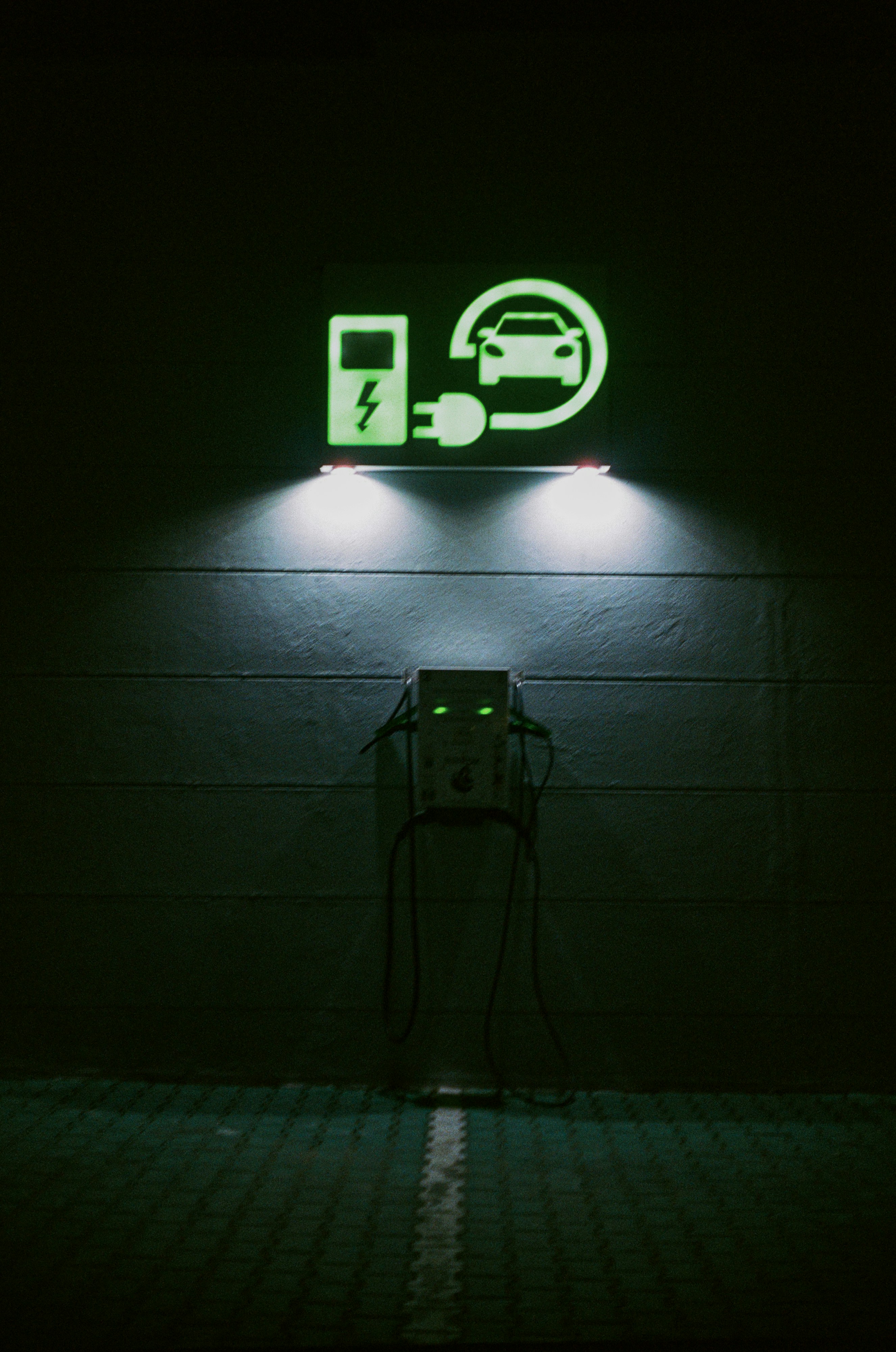 Neon green charging station for electric vehicles, featuring a battery icon and car silhouette, illuminated against a dark wall.