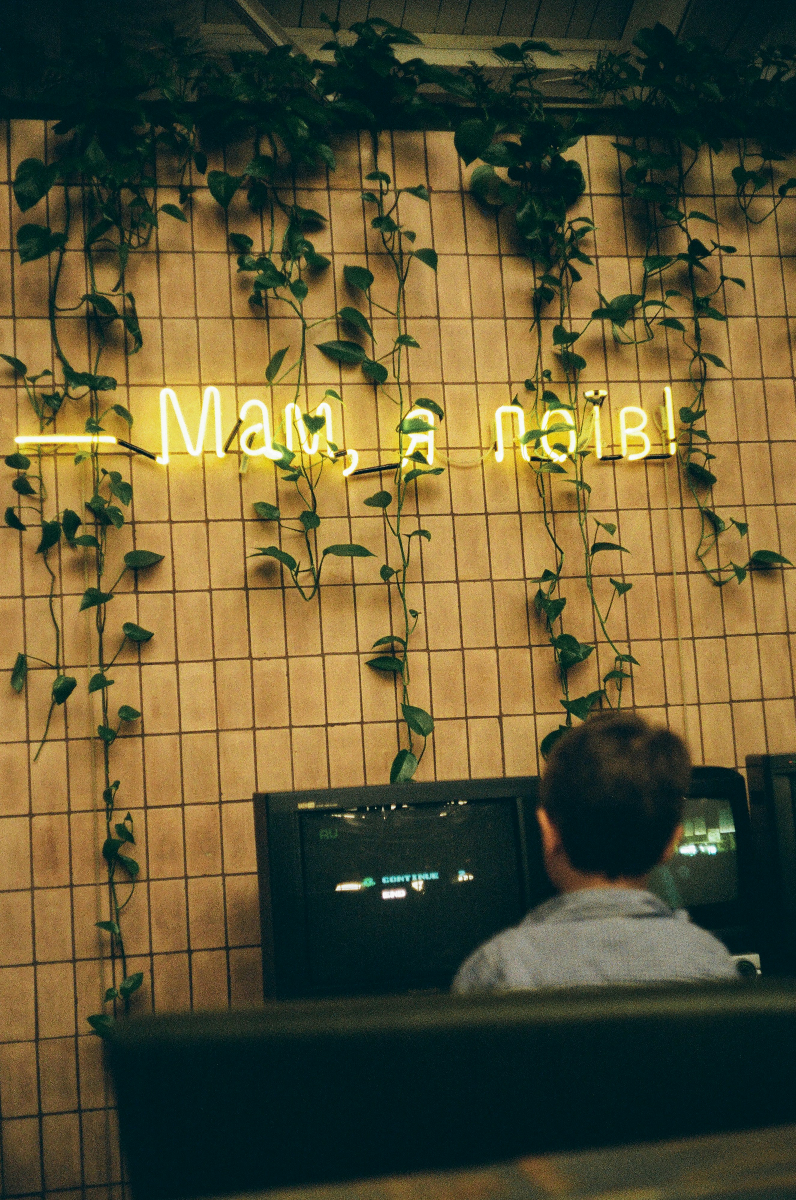 Ivy-clad tiled wall with a glowing neon sign casts warm light on a man facing two vintage CRT monitors. The scene evokes a late-night arcade vibe.
