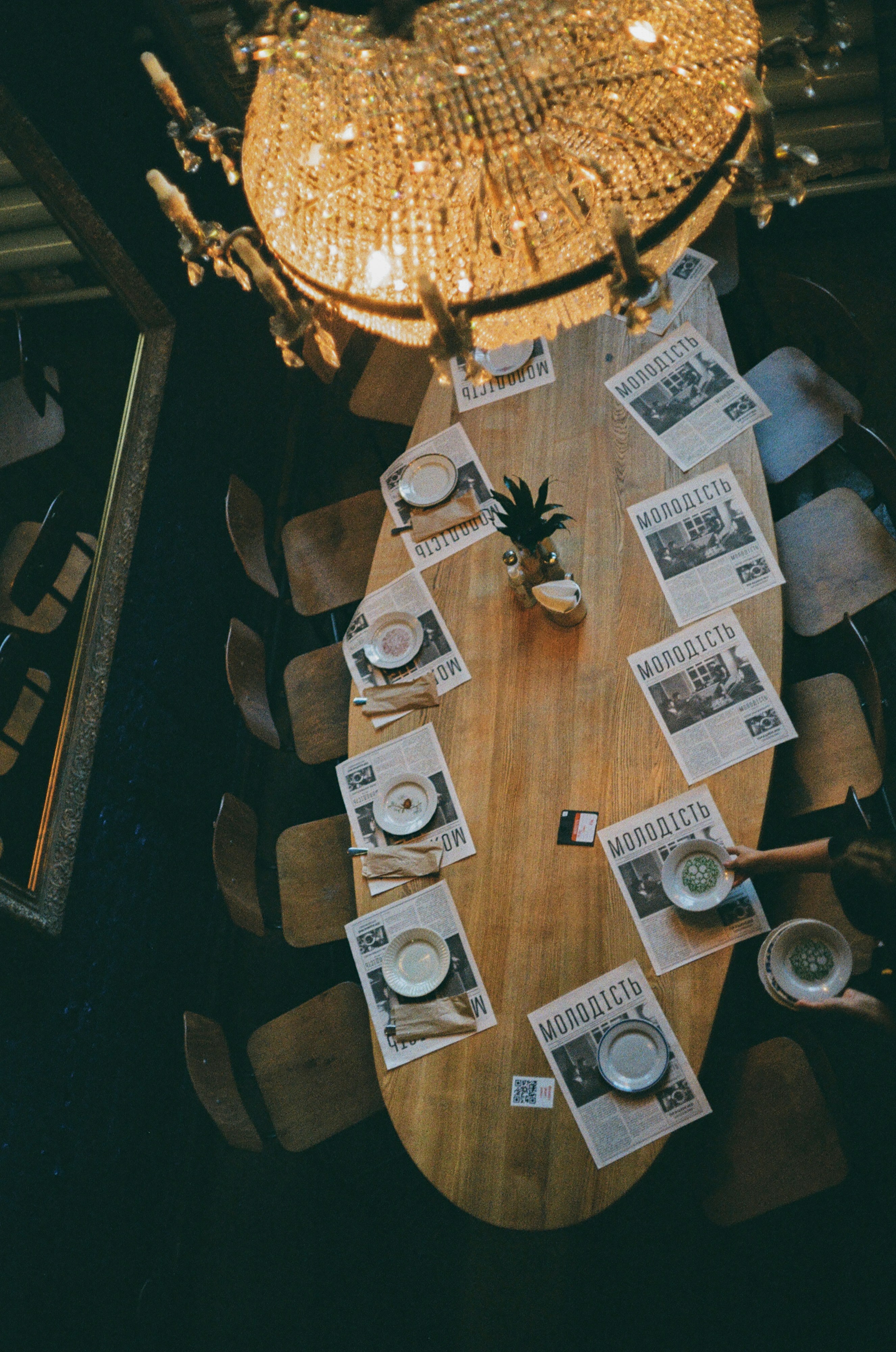 Aerial view of a wooden dining table set for a meal, adorned with newspapers and plates, beneath an elegant chandelier.