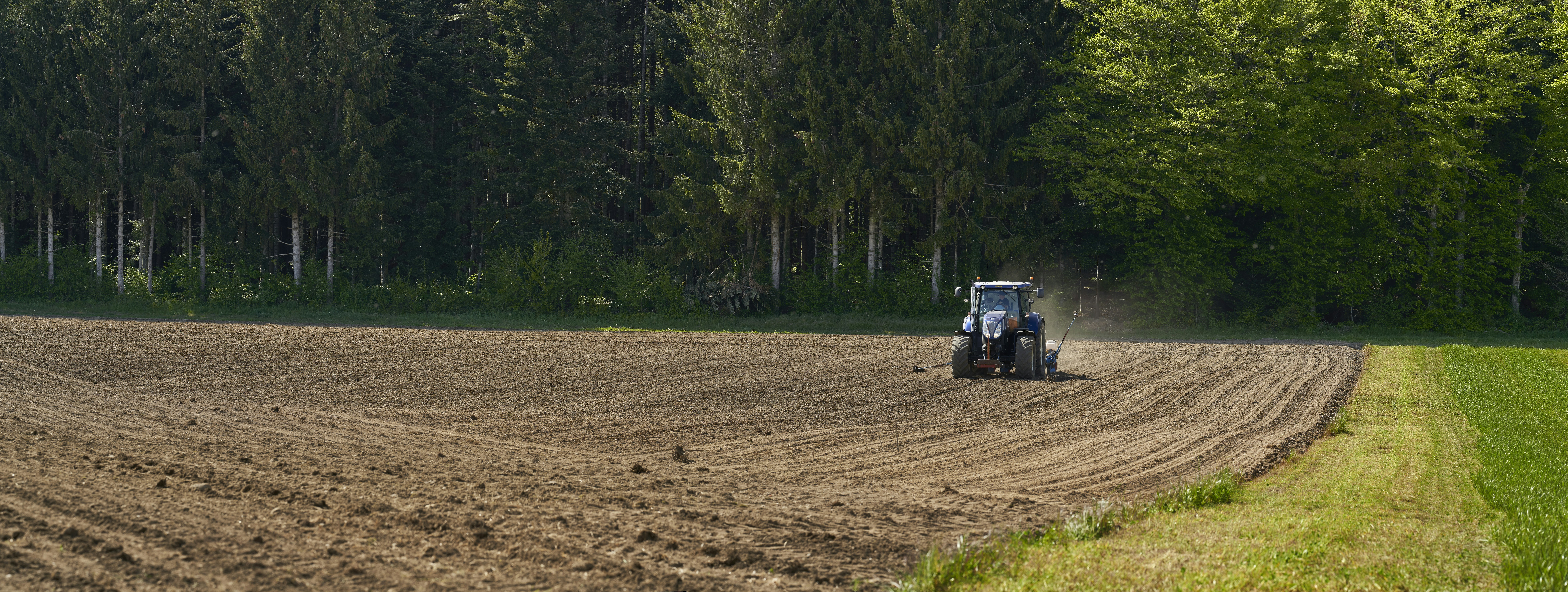 Tractor plowing a vast, freshly tilled field bordered by lush green trees.