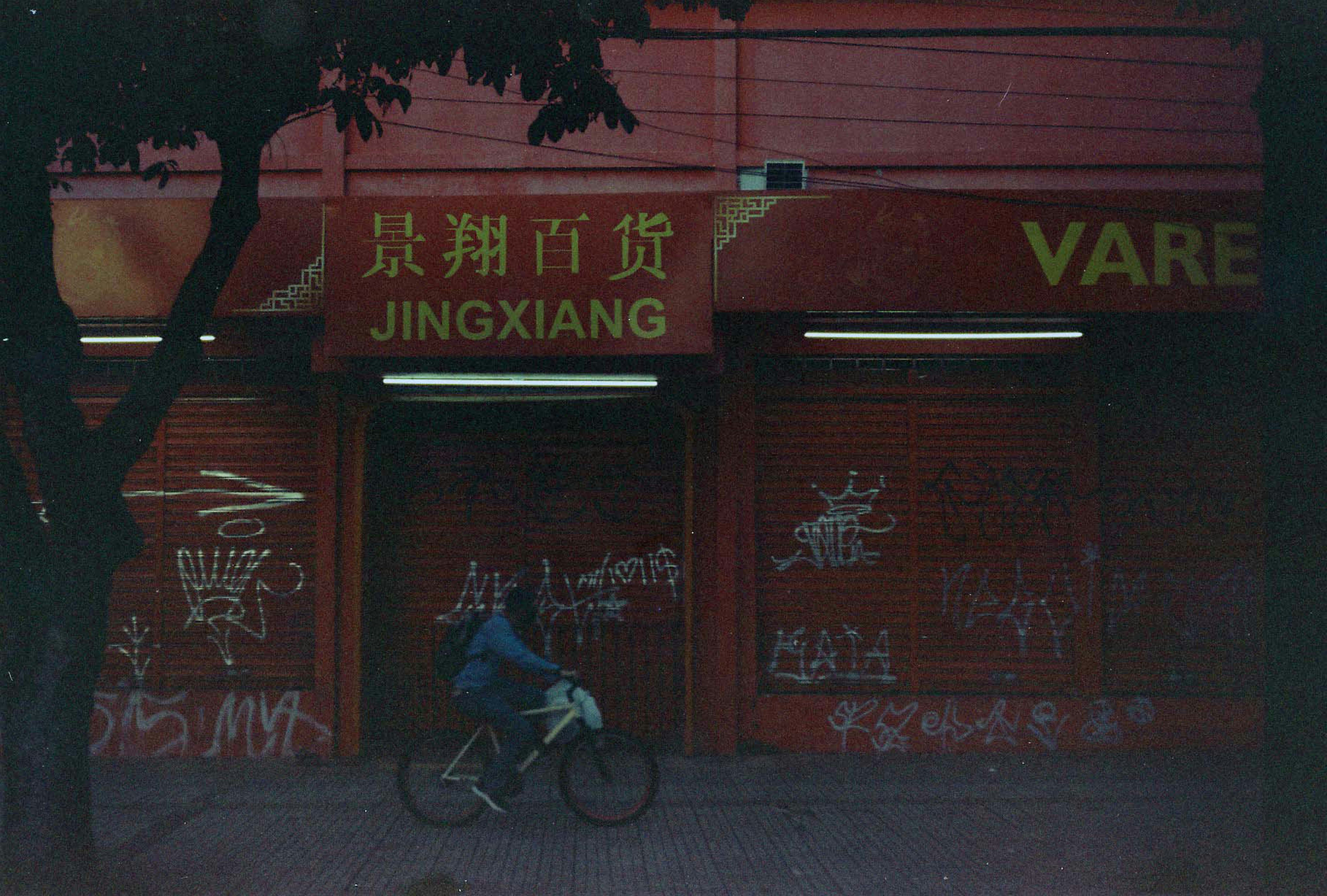 A cyclist rides past vibrant red storefronts adorned with graffiti, capturing the essence of urban life and artistic expression.