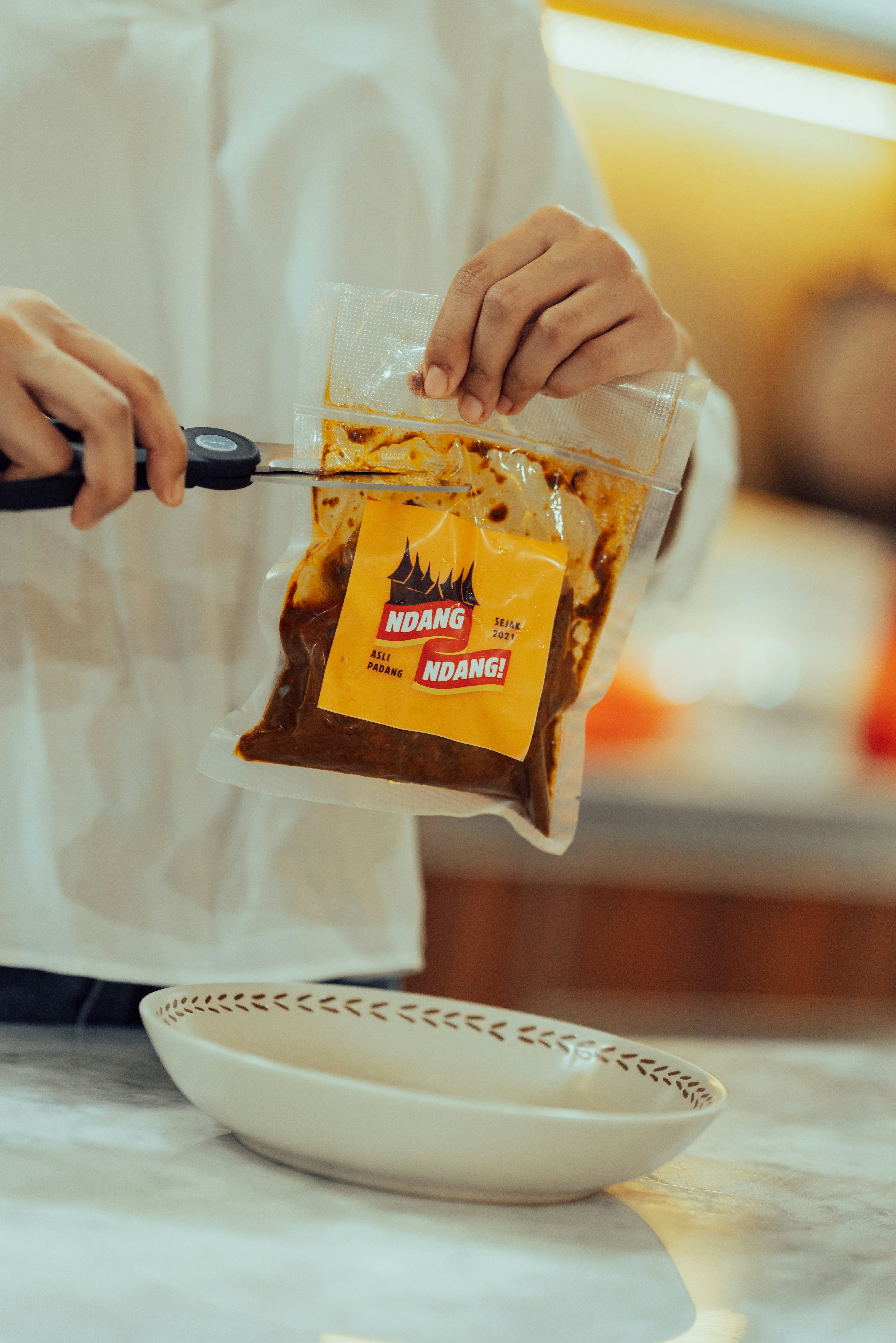 A hand holding a packet of Ndang! sauce, poised to pour its rich contents into a bowl. The kitchen setting provides a warm and inviting atmosphere.