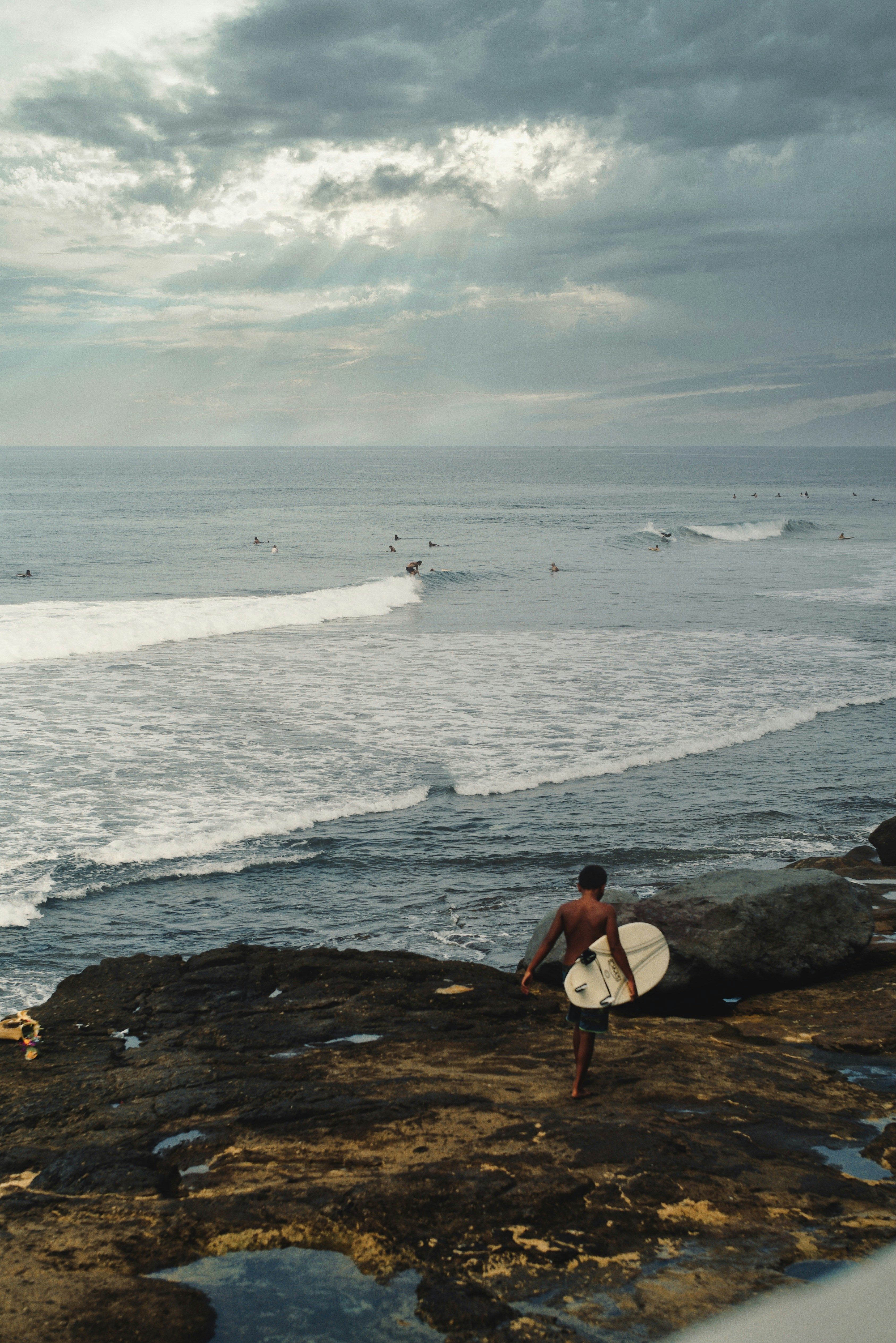 Surfer walking along rocky shoreline towards the ocean, with waves gently crashing in the background. Sunlight breaks through clouds, illuminating the scene.